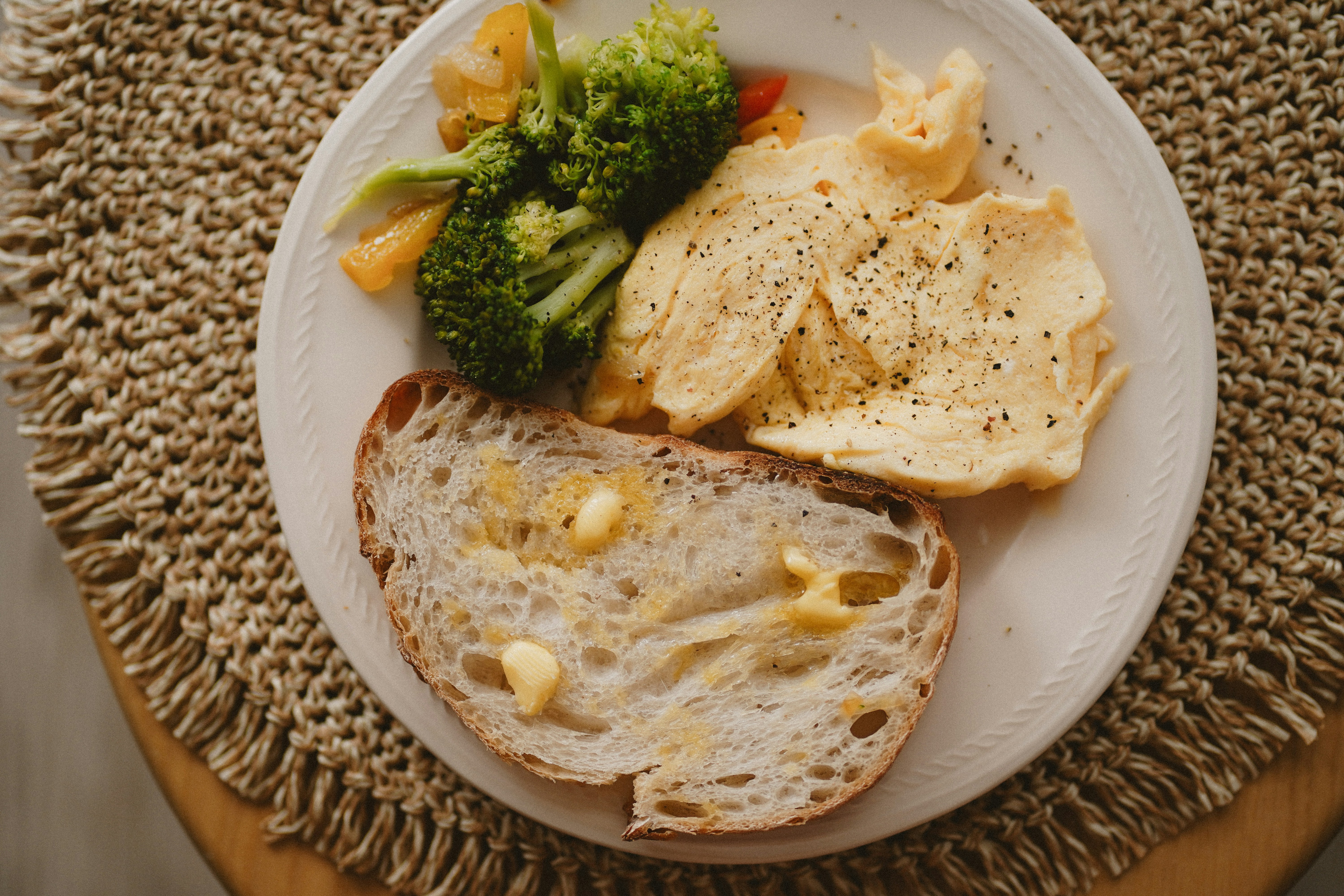 Top-down view of a comforting breakfast plate featuring fluffy scrambled eggs, steamed vegetables, and rustic bread with melted butter. | A plate of food with toast, chicken, and vegetables.