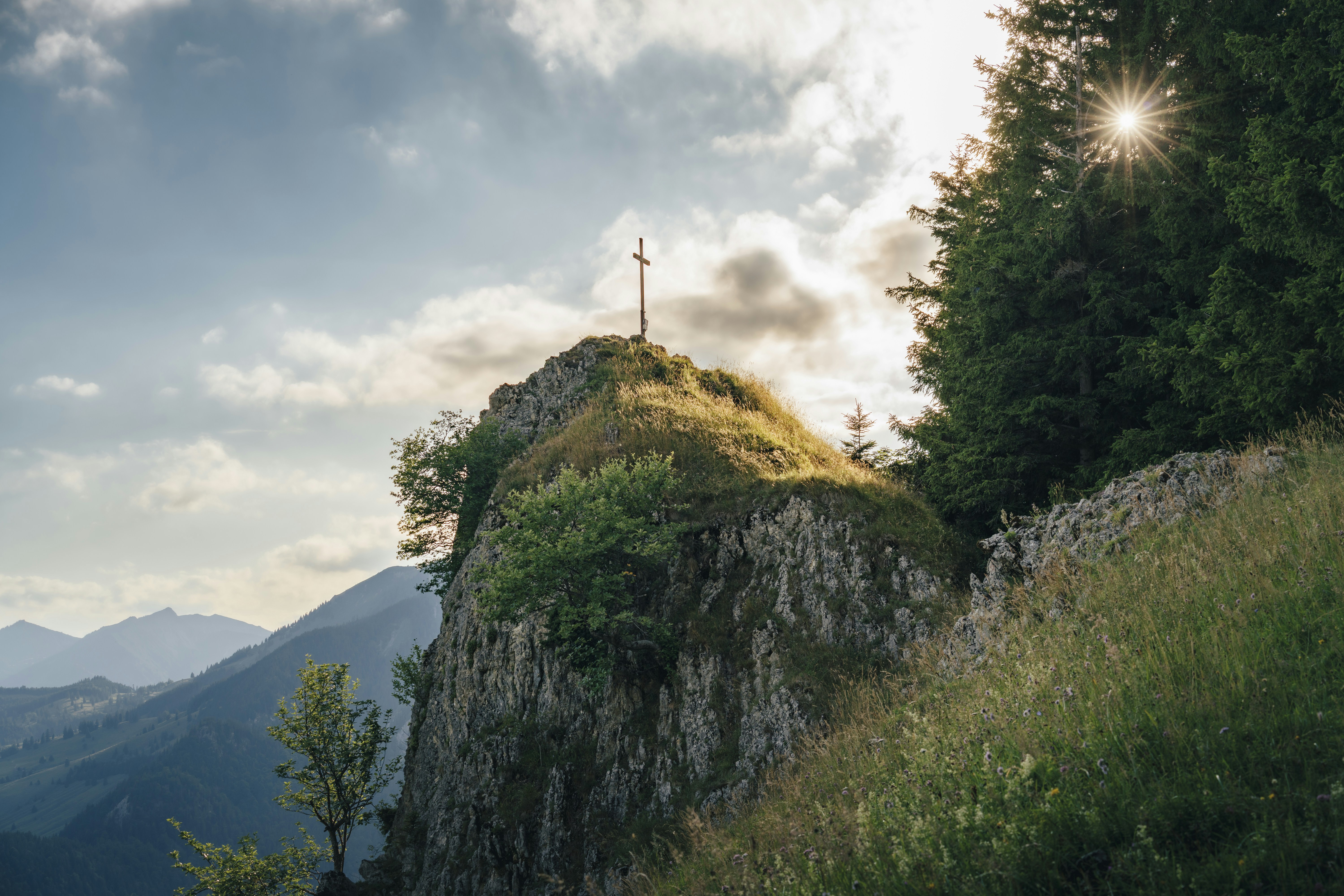 A solitary cross stands atop a rocky outcrop, surrounded by lush greenery and distant mountains under a partly cloudy sky.