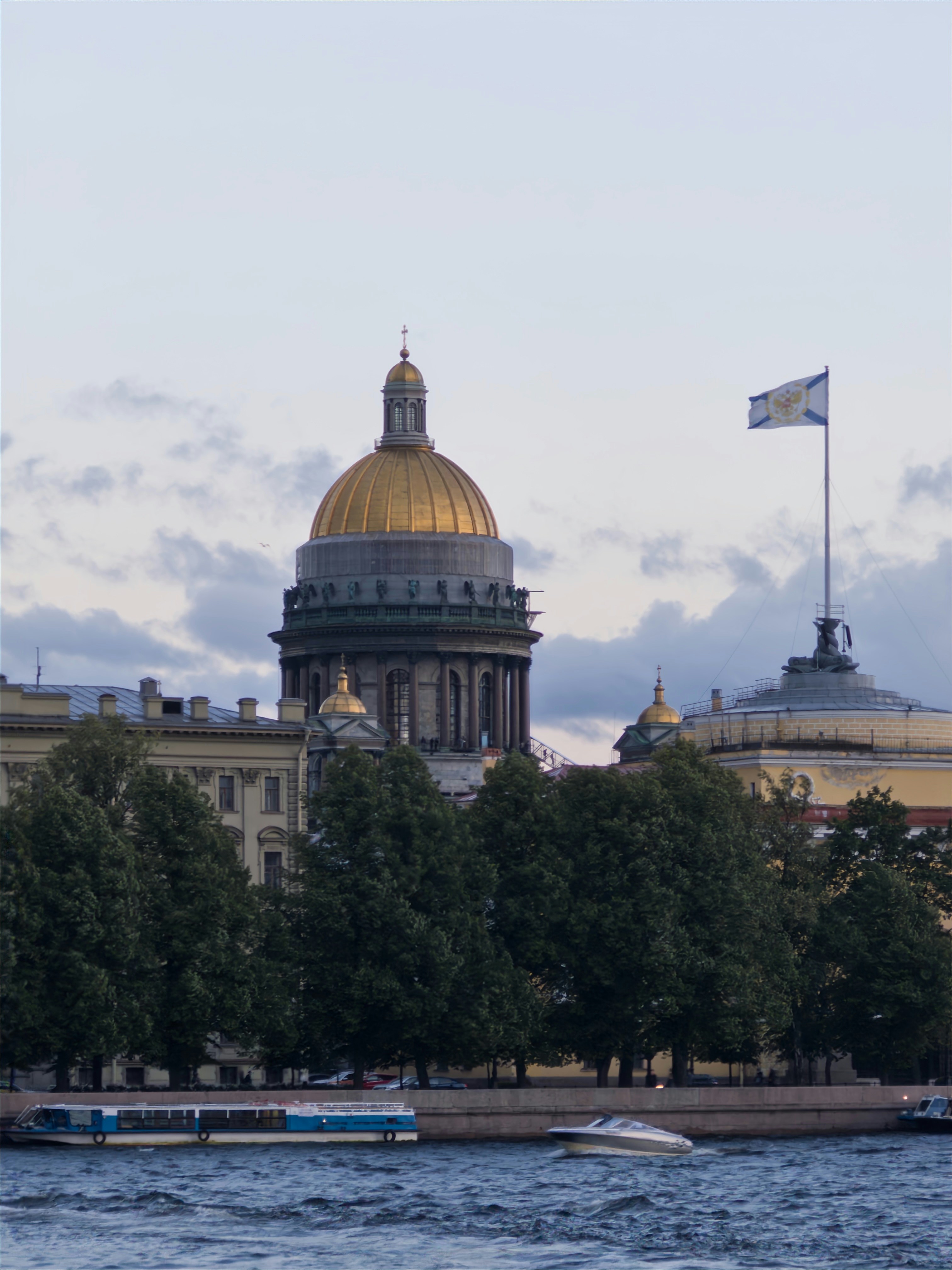Historic dome of a prominent building rises above lush trees along a riverbank, framed by a cloudy sky. A flag flutters in the breeze.