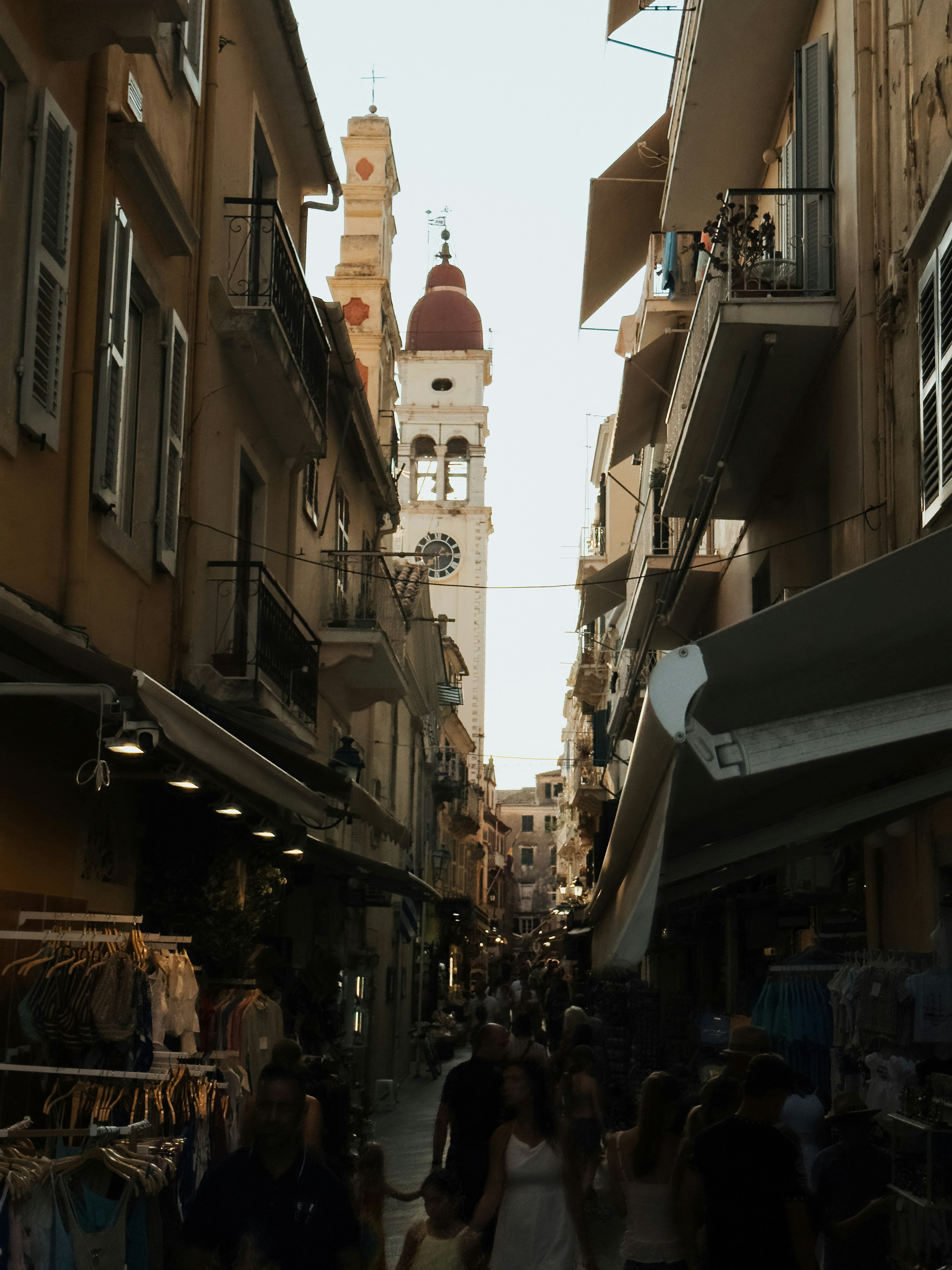 Narrow street in Corfu Old Town leading to the iconic church clock tower. | Narrow european street with shops and bell tower