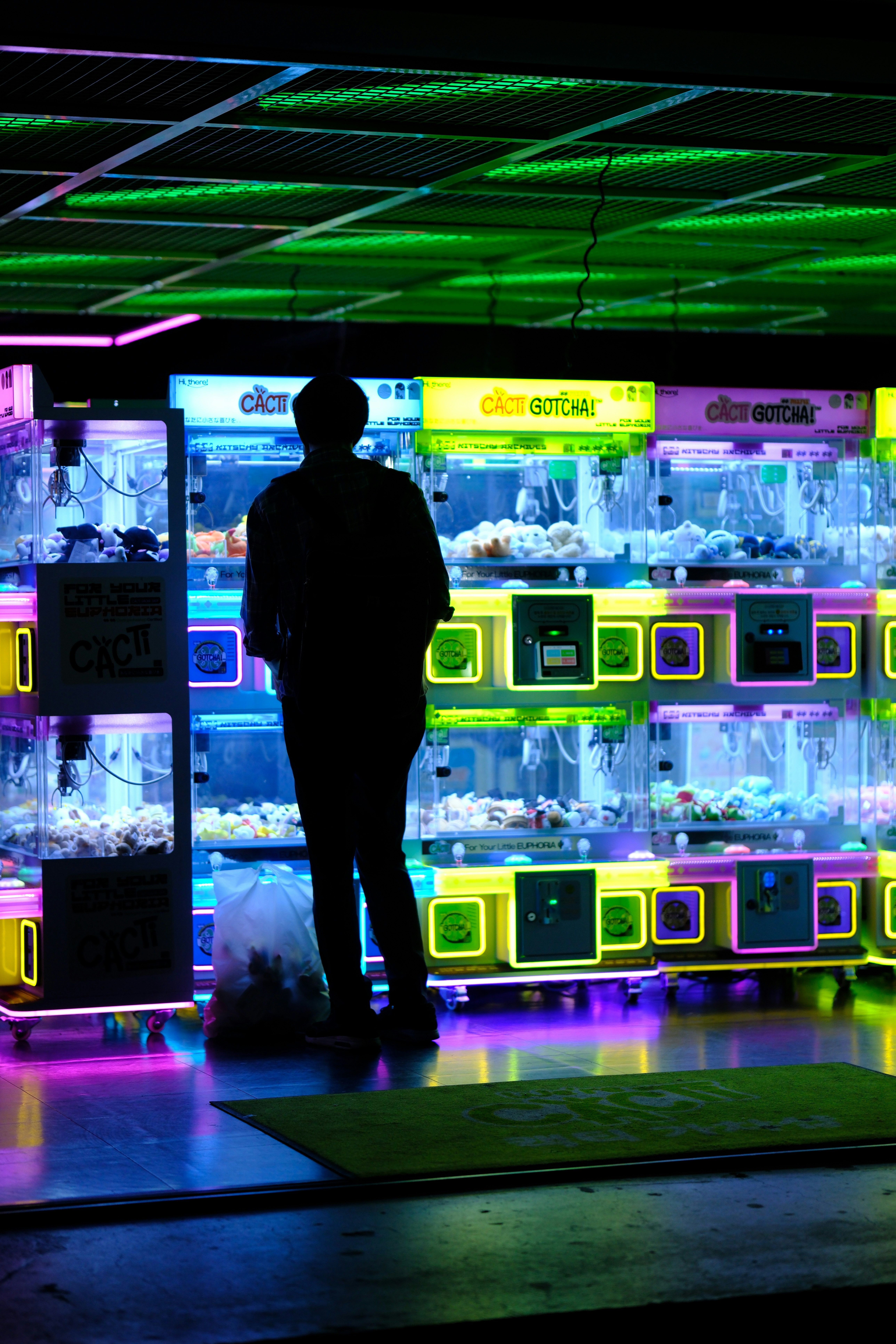 Silhouette of a person standing in front of brightly lit claw machines filled with colorful plush toys in a vibrant arcade setting.