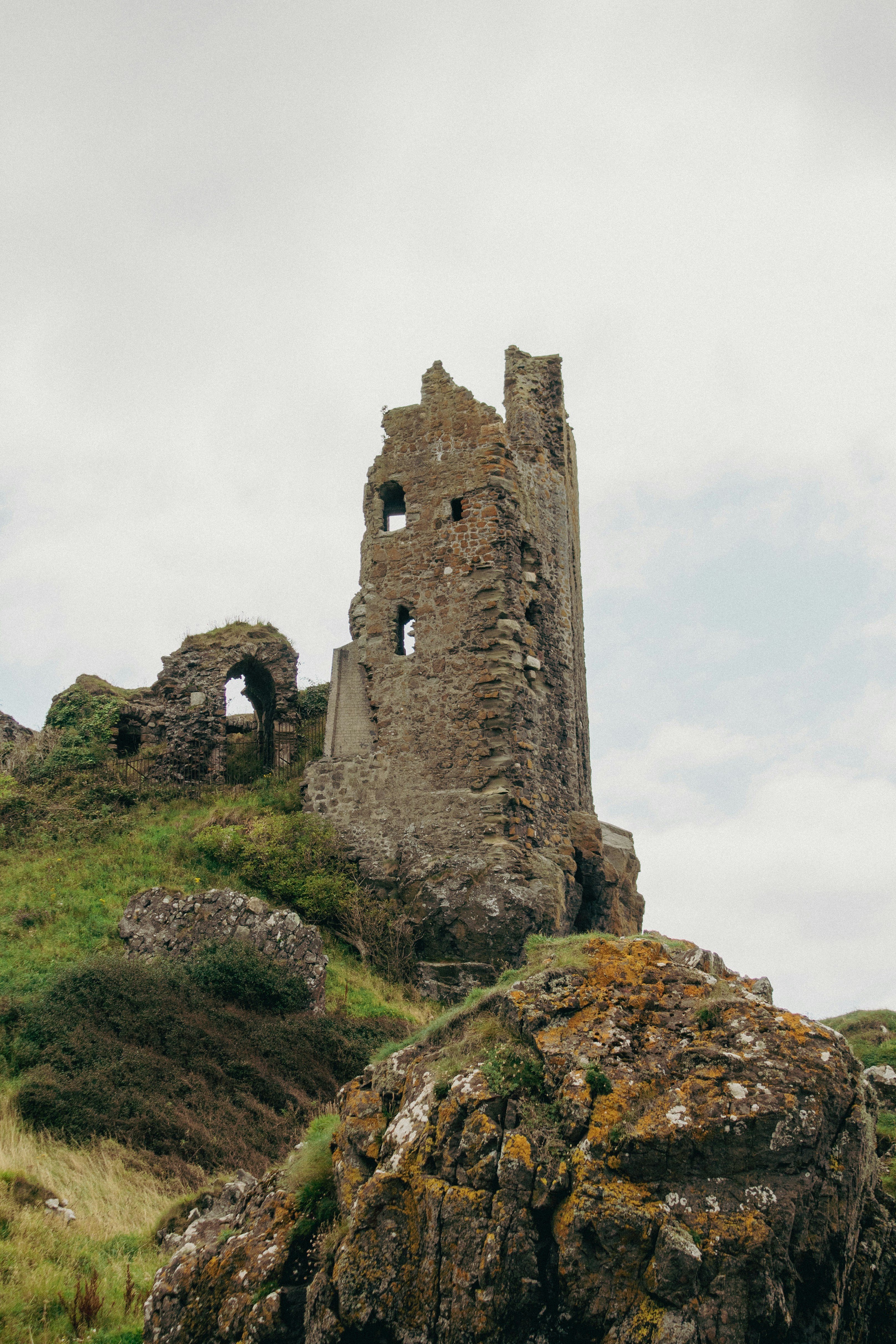 Ancient stone tower ruins rise from a grassy hillside, framed by rocky outcrops and a cloudy sky.