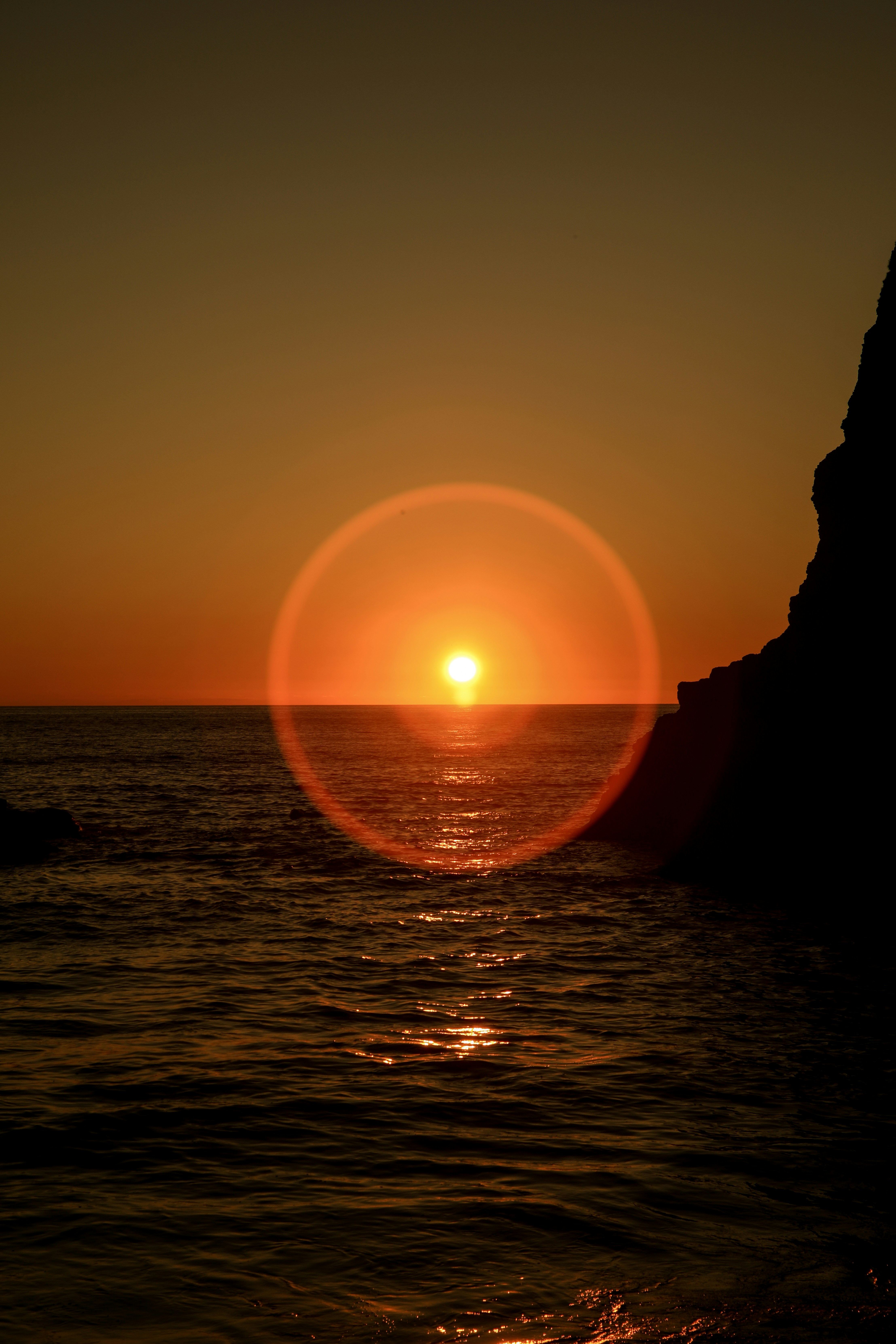 Vibrant sunset illuminating the ocean with a lens flare, framed by rocky silhouettes on the right.