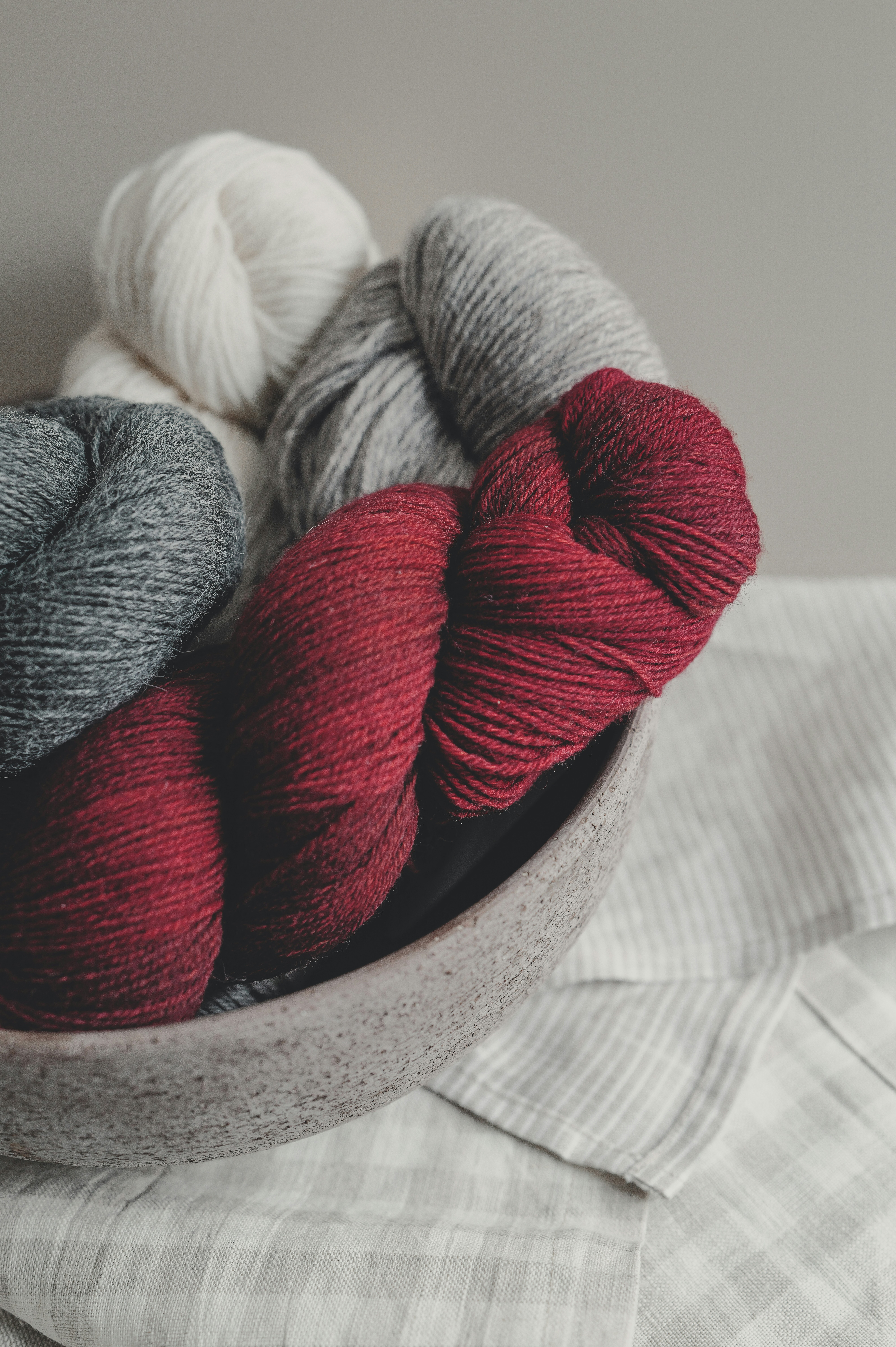 Skeins of yarn in red, gray, and white are arranged in a stone bowl. The soft texture and muted colors create a calm and cozy feeling. | Bowl of red, grey, and white yarn