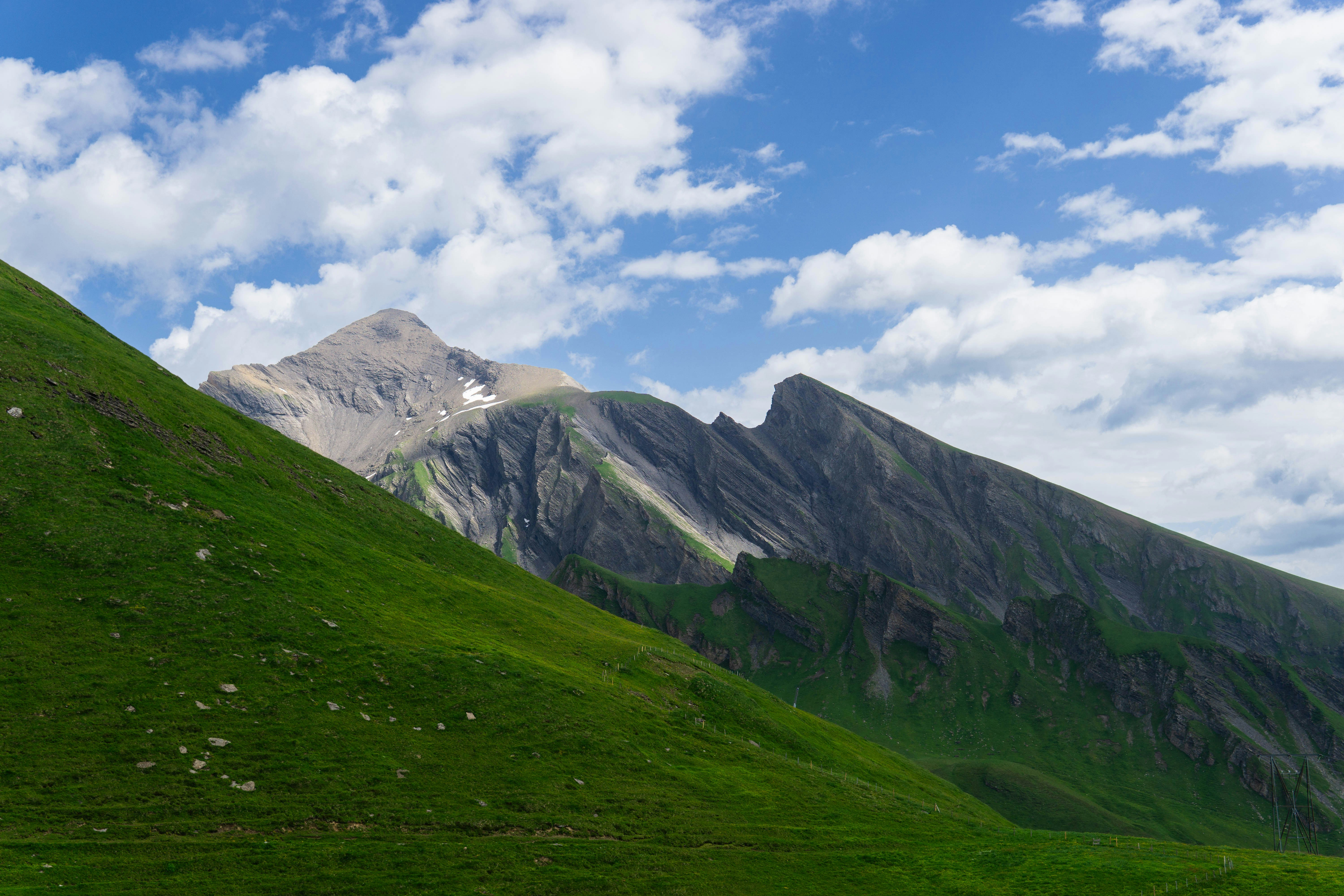 Majestic peaks rise above lush green hills under a dynamic sky, showcasing the beauty of nature's rugged terrain.