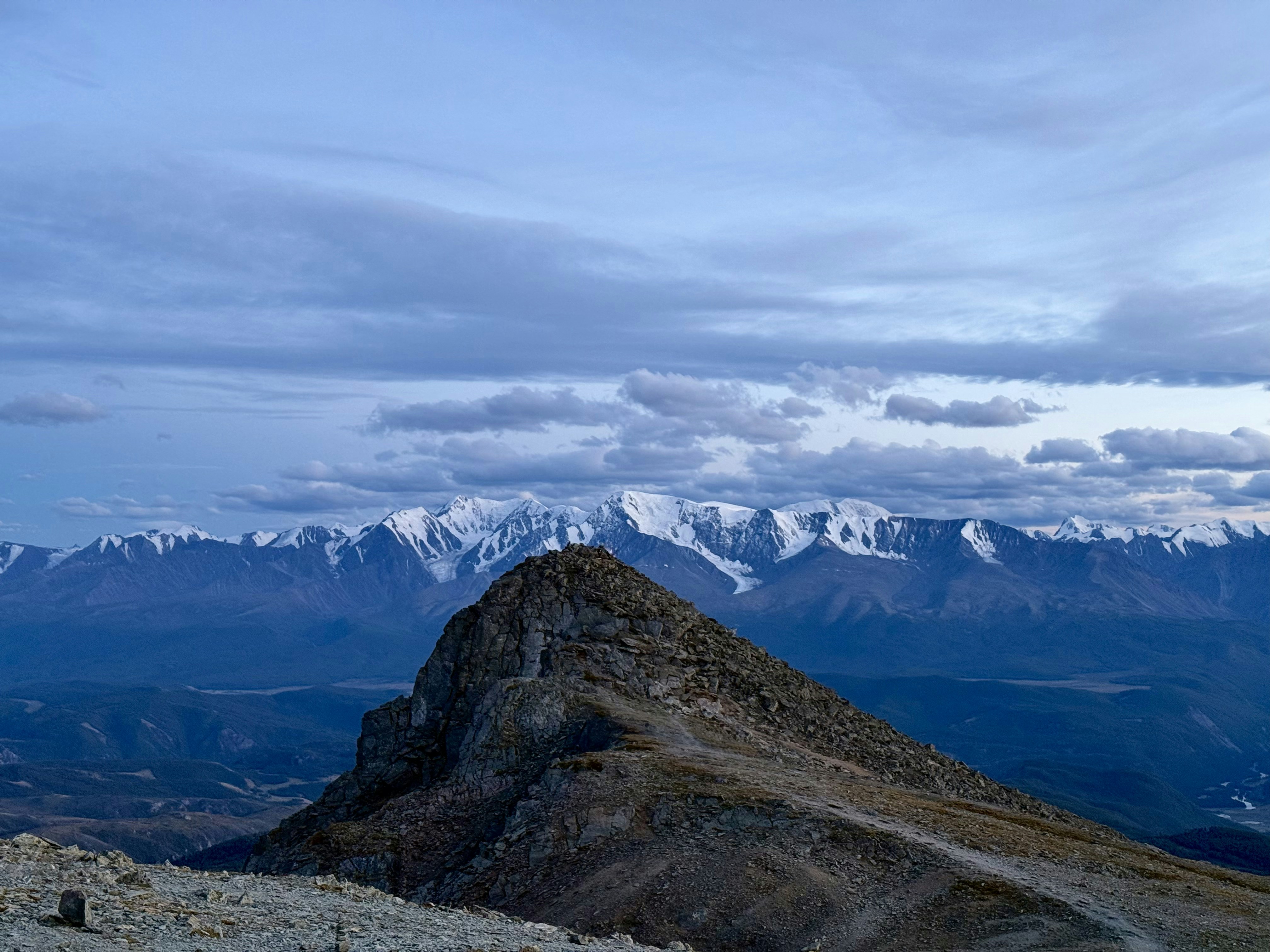 Rocky mountain ridge with snow-capped peaks in the background under a cloudy sky.