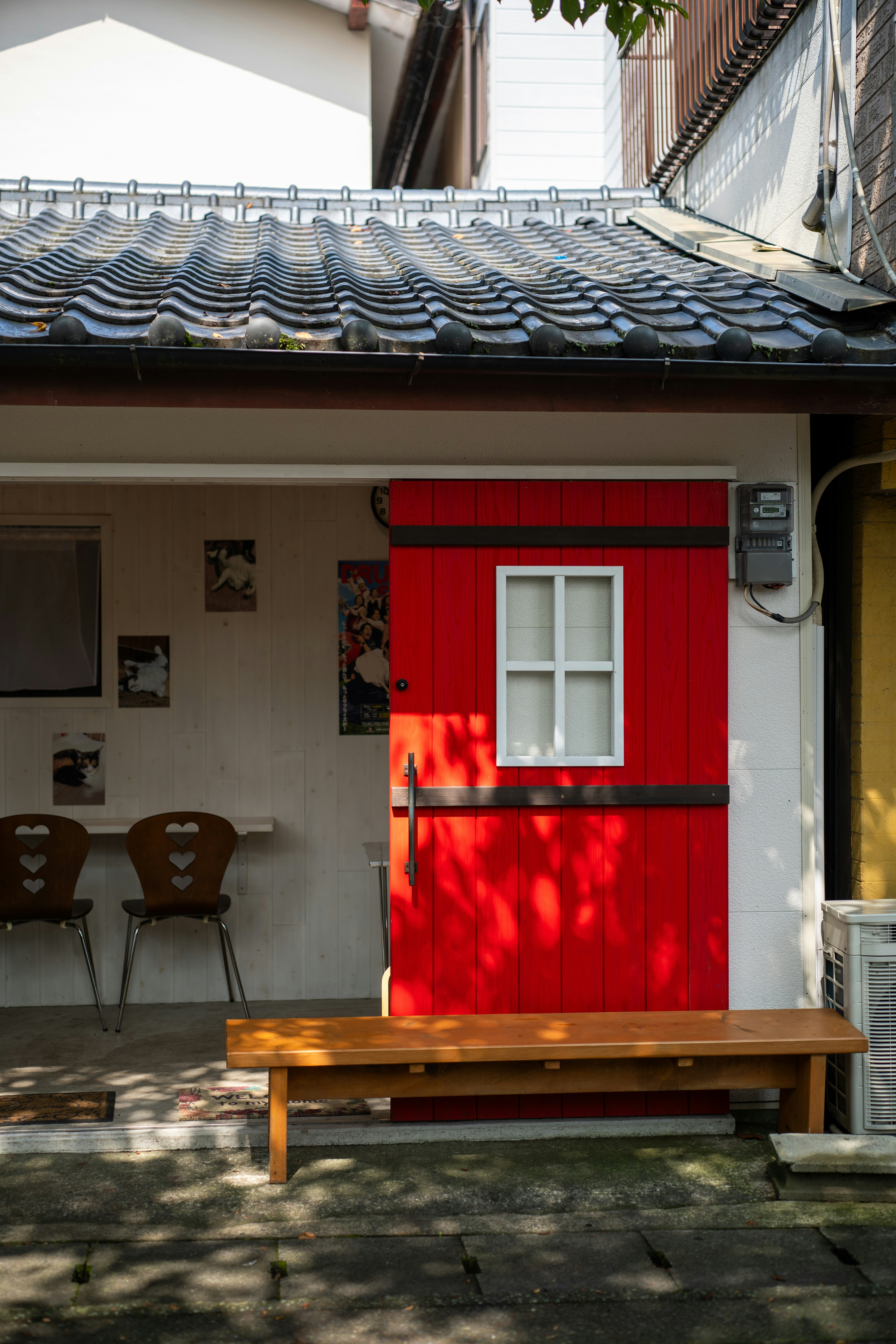 A bright red door with a white window stands prominently against a softly lit wall, accompanied by wooden chairs and a bench, creating an inviting atmosphere.