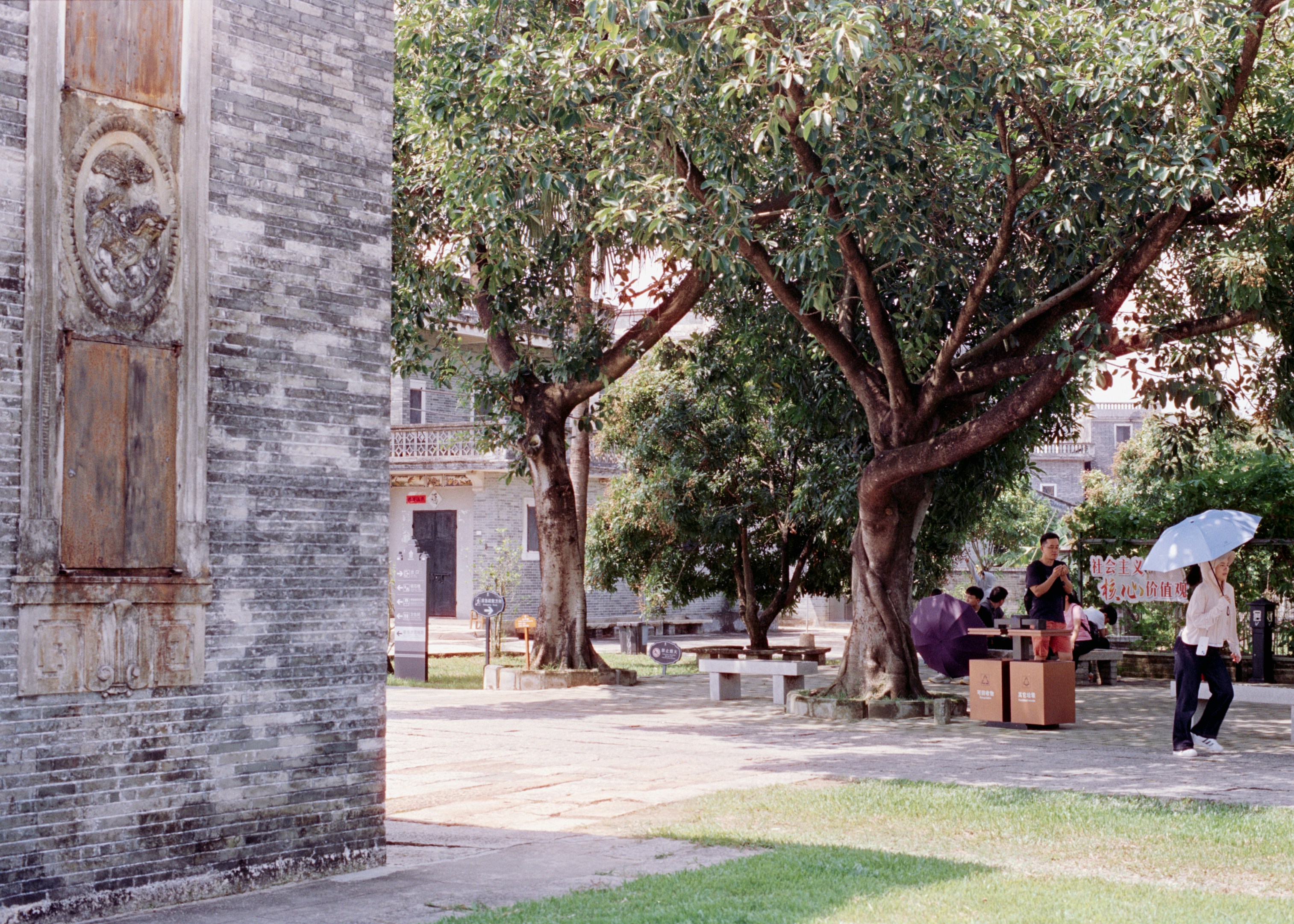 People gather under trees in a courtyard with brick building. photo ...