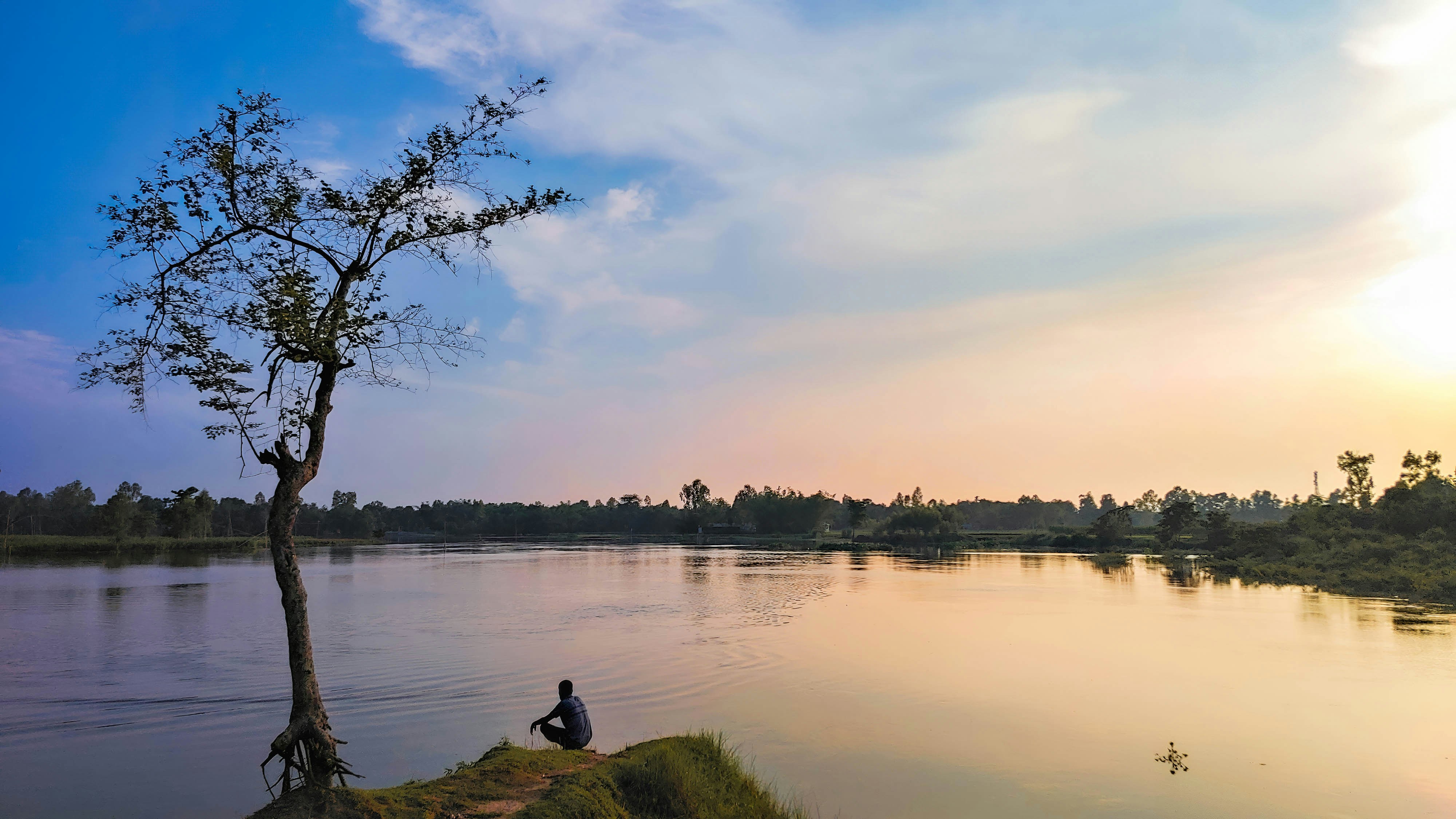 A lone figure sits beside a tranquil river, framed by a solitary tree under a colorful sky at sunset.