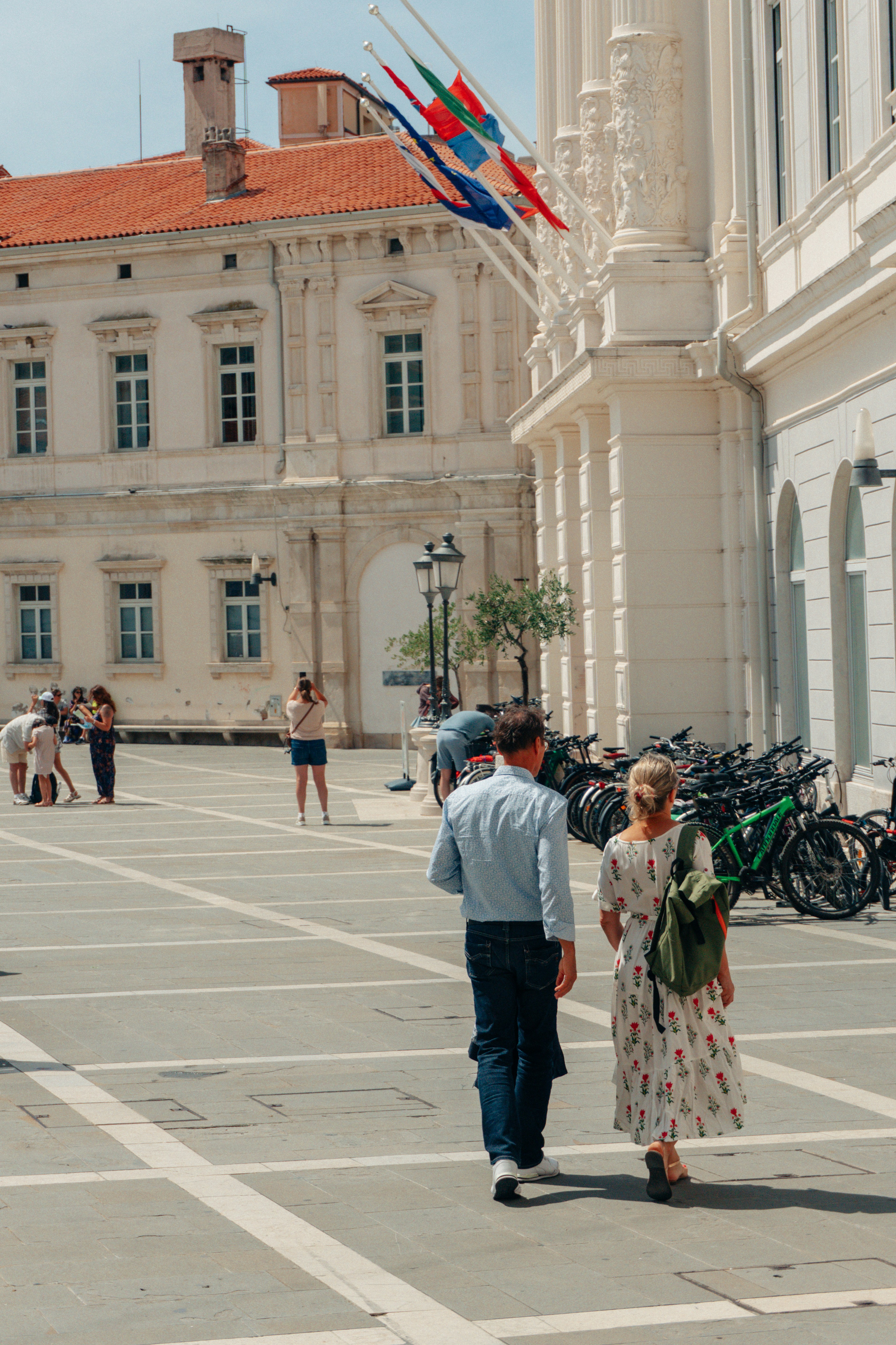 Couple walking hand-in-hand along a sunlit plaza, surrounded by historic architecture and bicycles parked nearby.