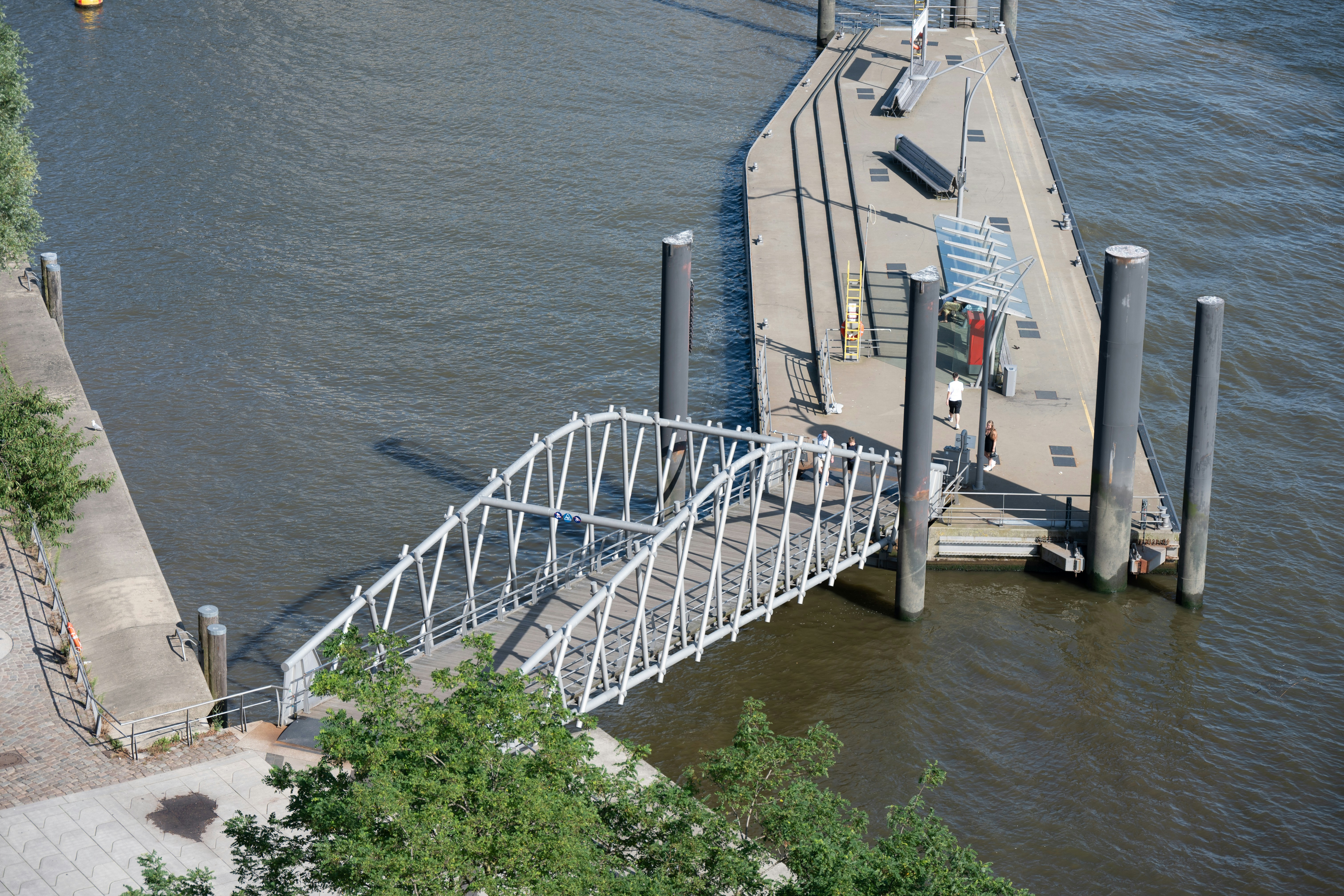 A modern pedestrian bridge connects to a dock over calm waters, surrounded by lush greenery and urban architecture.