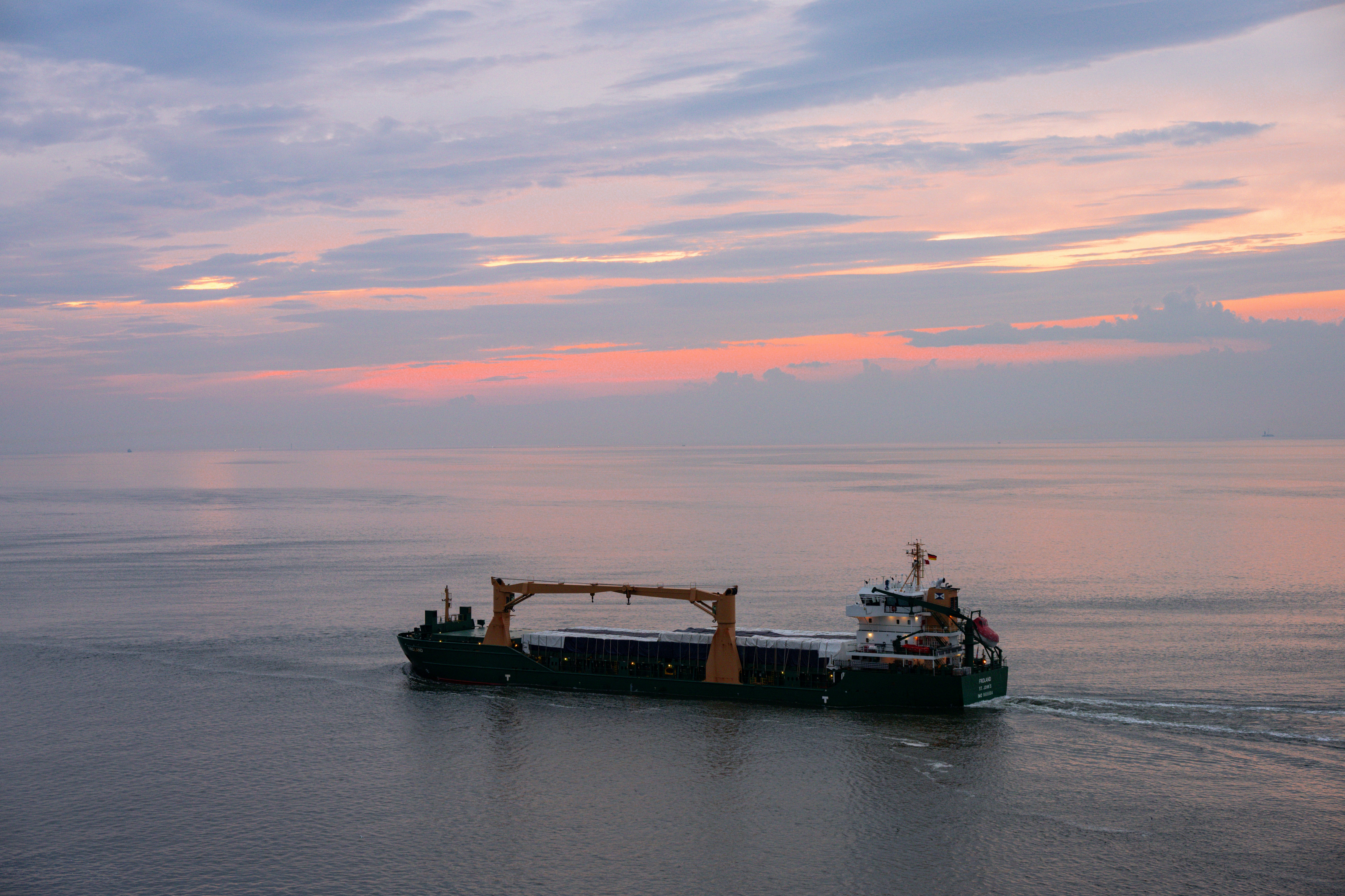 Cargo ship gliding through calm waters under a pastel sky at sunrise.