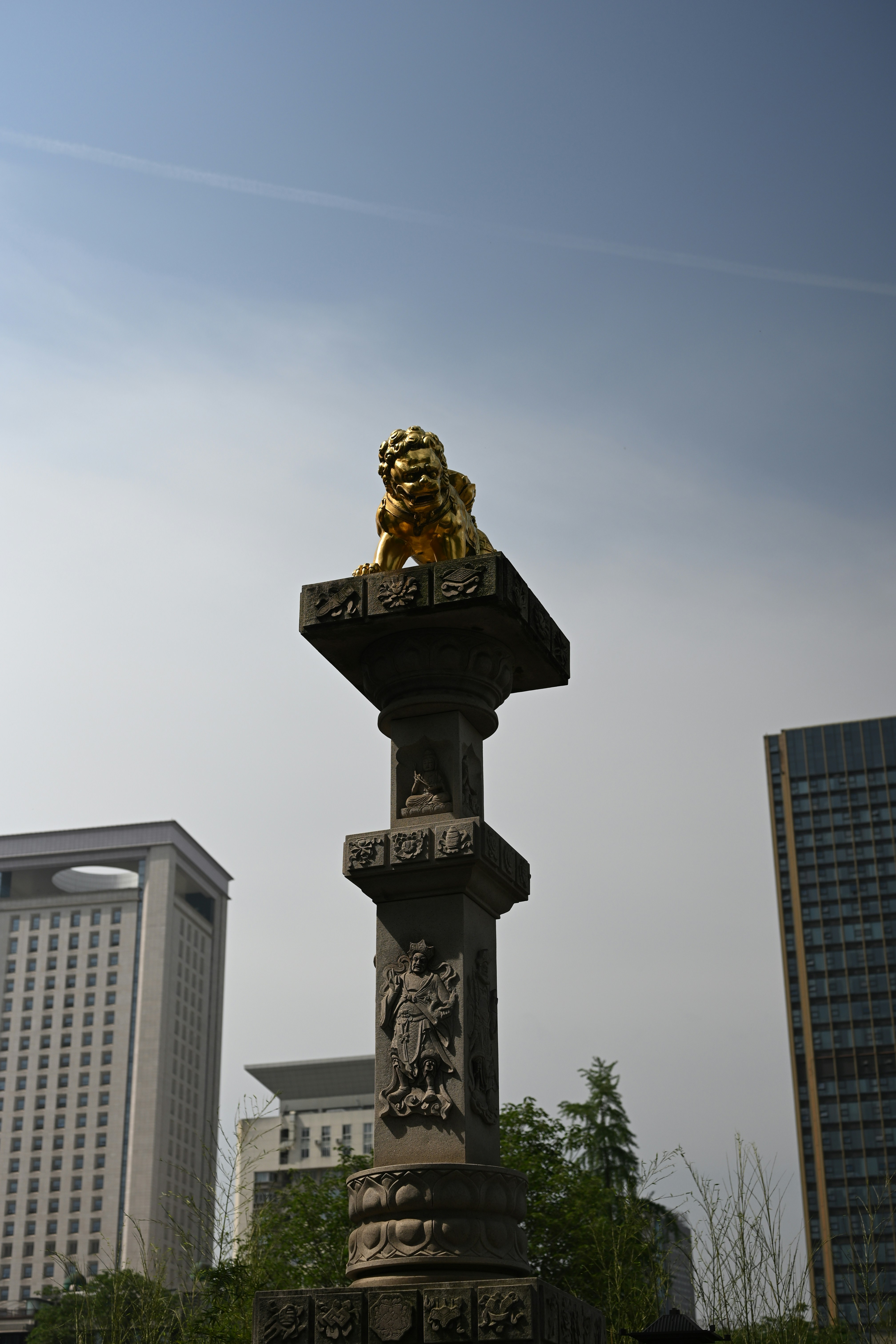 A golden lion statue perched atop a decorated pedestal, surrounded by modern buildings and greenery. The scene captures the contrast between tradition and contemporary architecture.