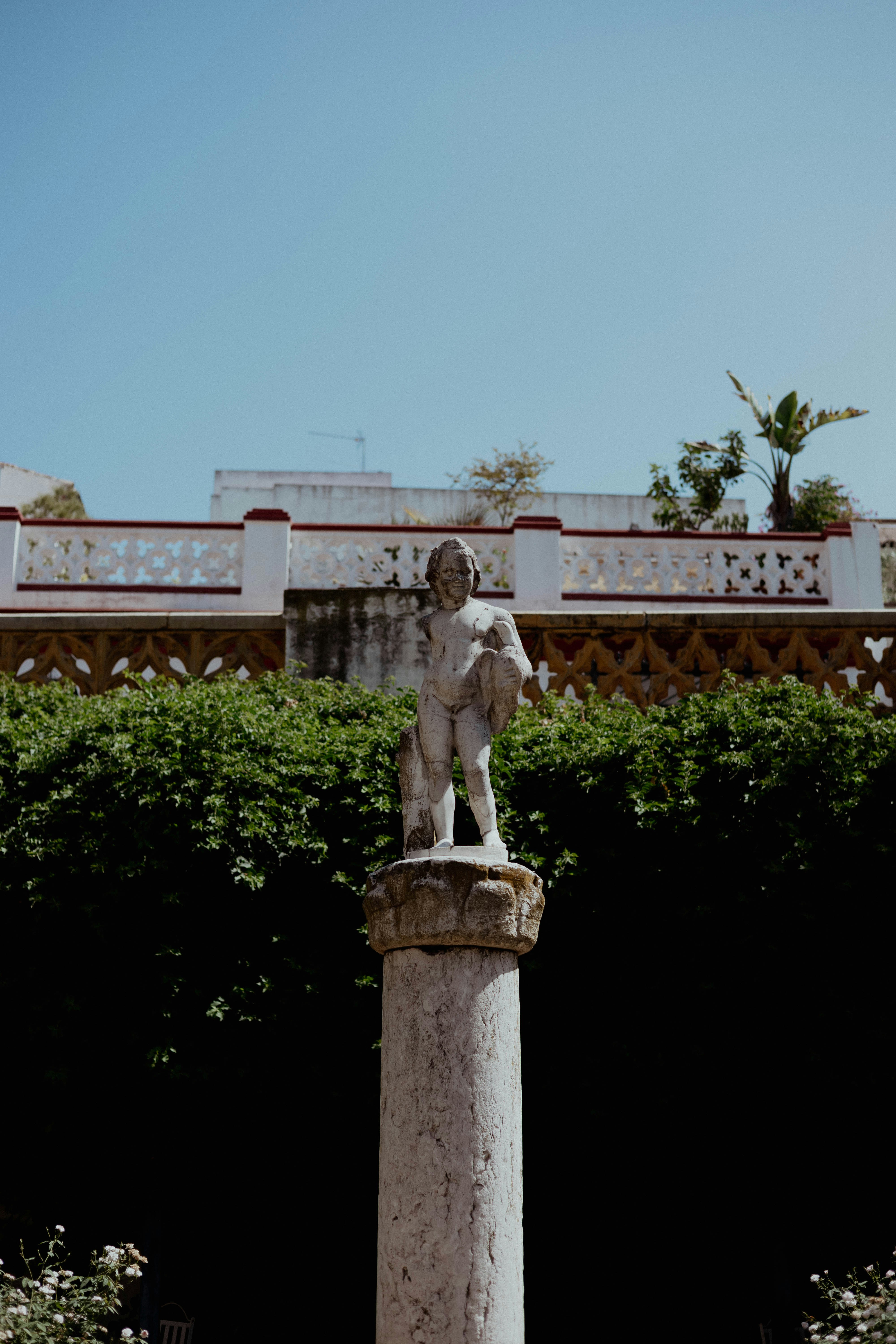 A weathered statue of a young boy stands atop a stone pedestal, surrounded by lush greenery and a clear blue sky.