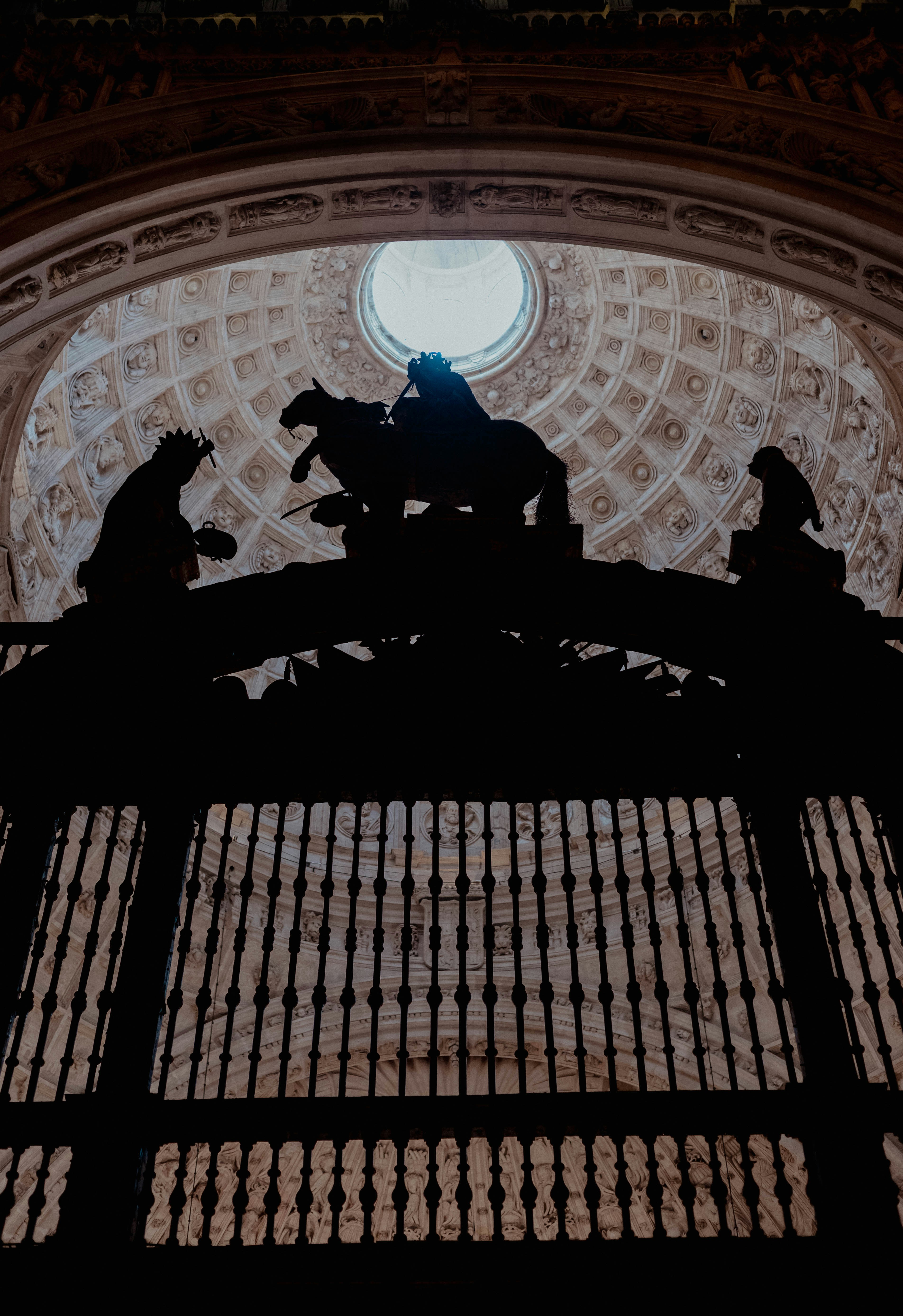 Silhouetted figures atop an ornate structure beneath a circular skylight, showcasing intricate architectural details. The interplay of light and shadow creates a dramatic effect.