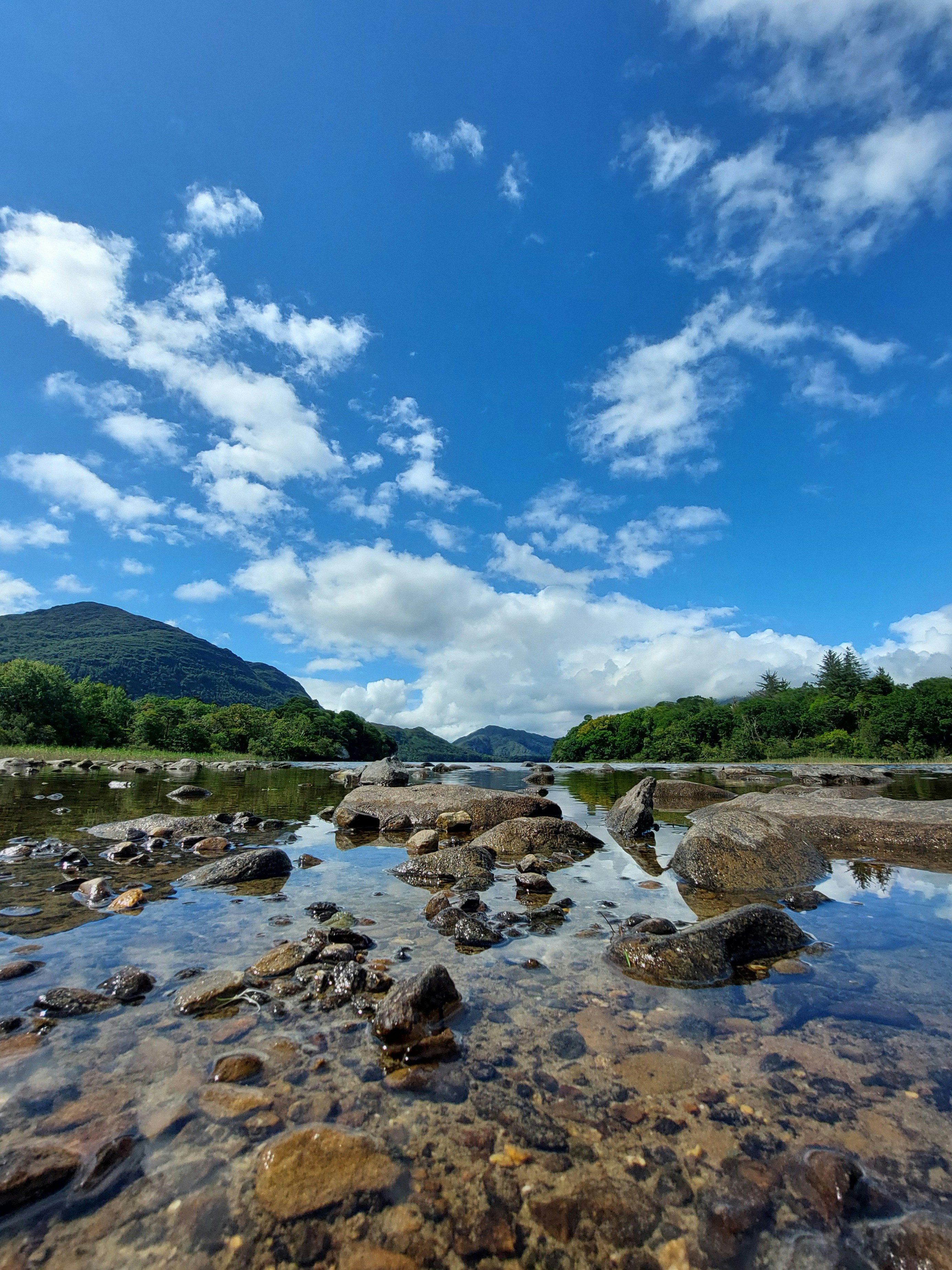 Clear riverbed with stones in the foreground, calm water reflecting the blue sky; lush green banks and distant hills create a tranquil landscape scene.