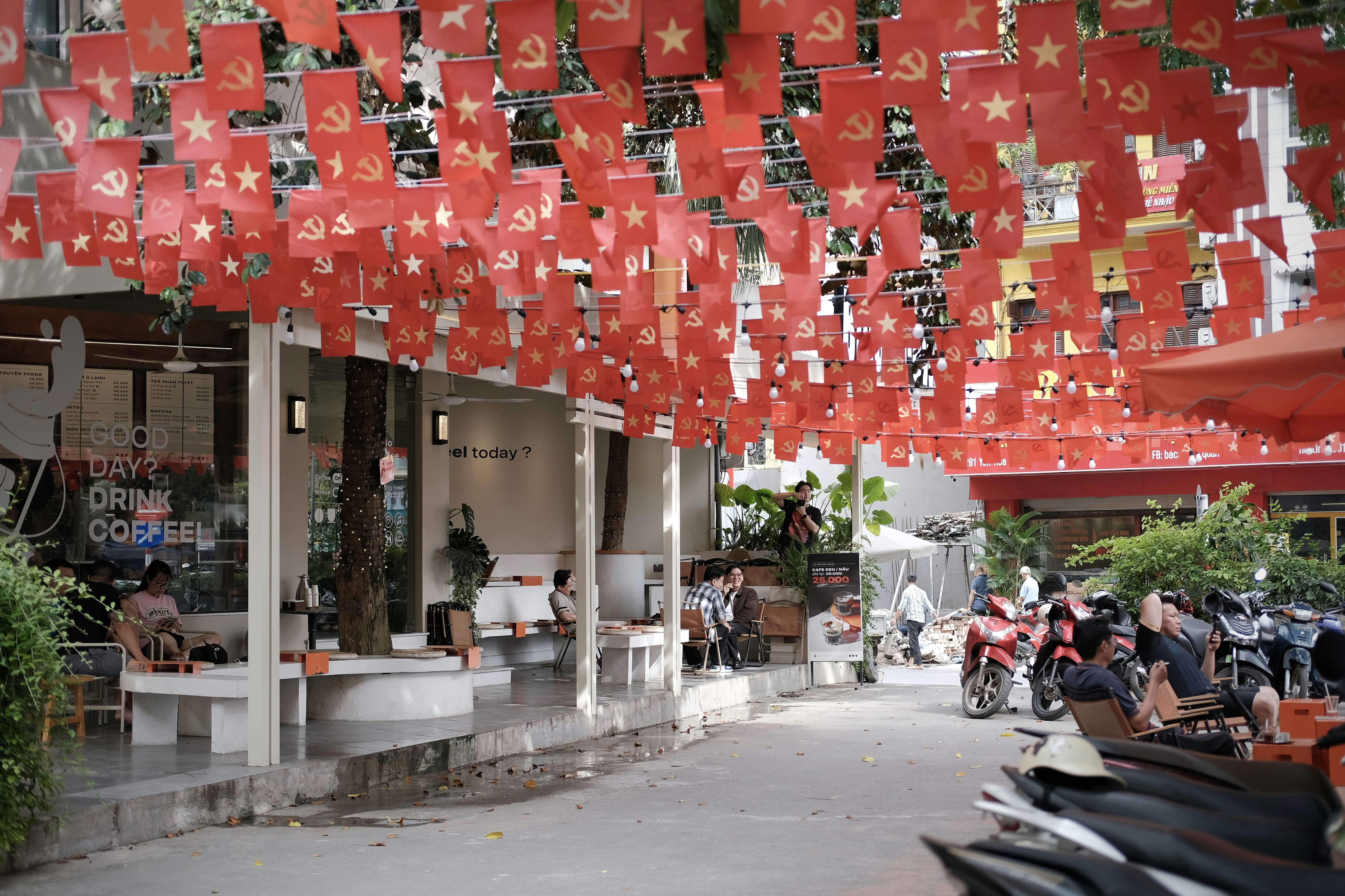 A vibrant display of red flags hangs above a bustling café, creating a lively atmosphere. Patrons enjoy their coffee beneath the colorful decorations.