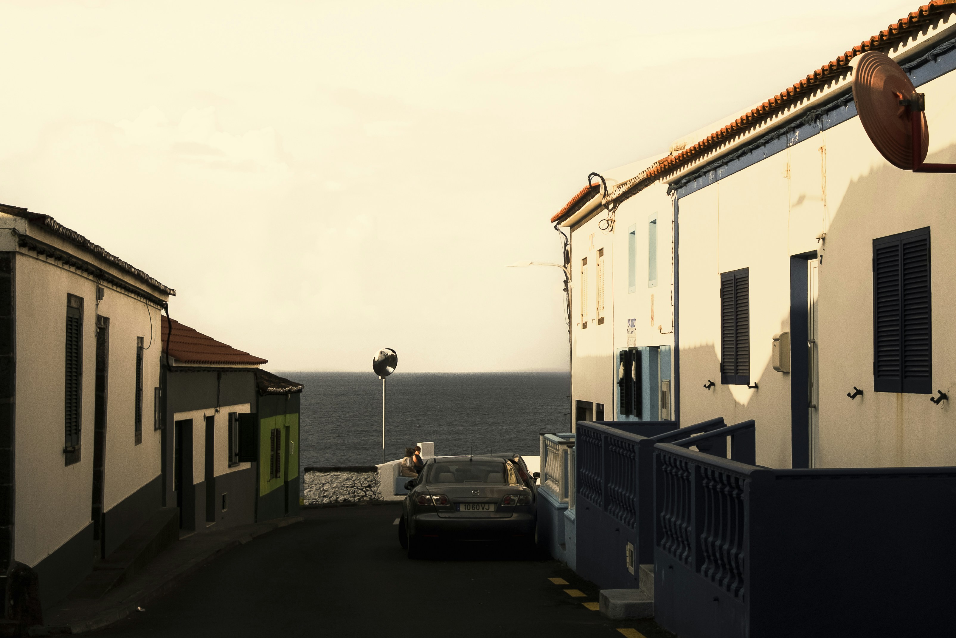 Narrow street lined with colorful houses leading to the ocean, showcasing a serene coastal atmosphere. A parked car adds to the tranquil scene.