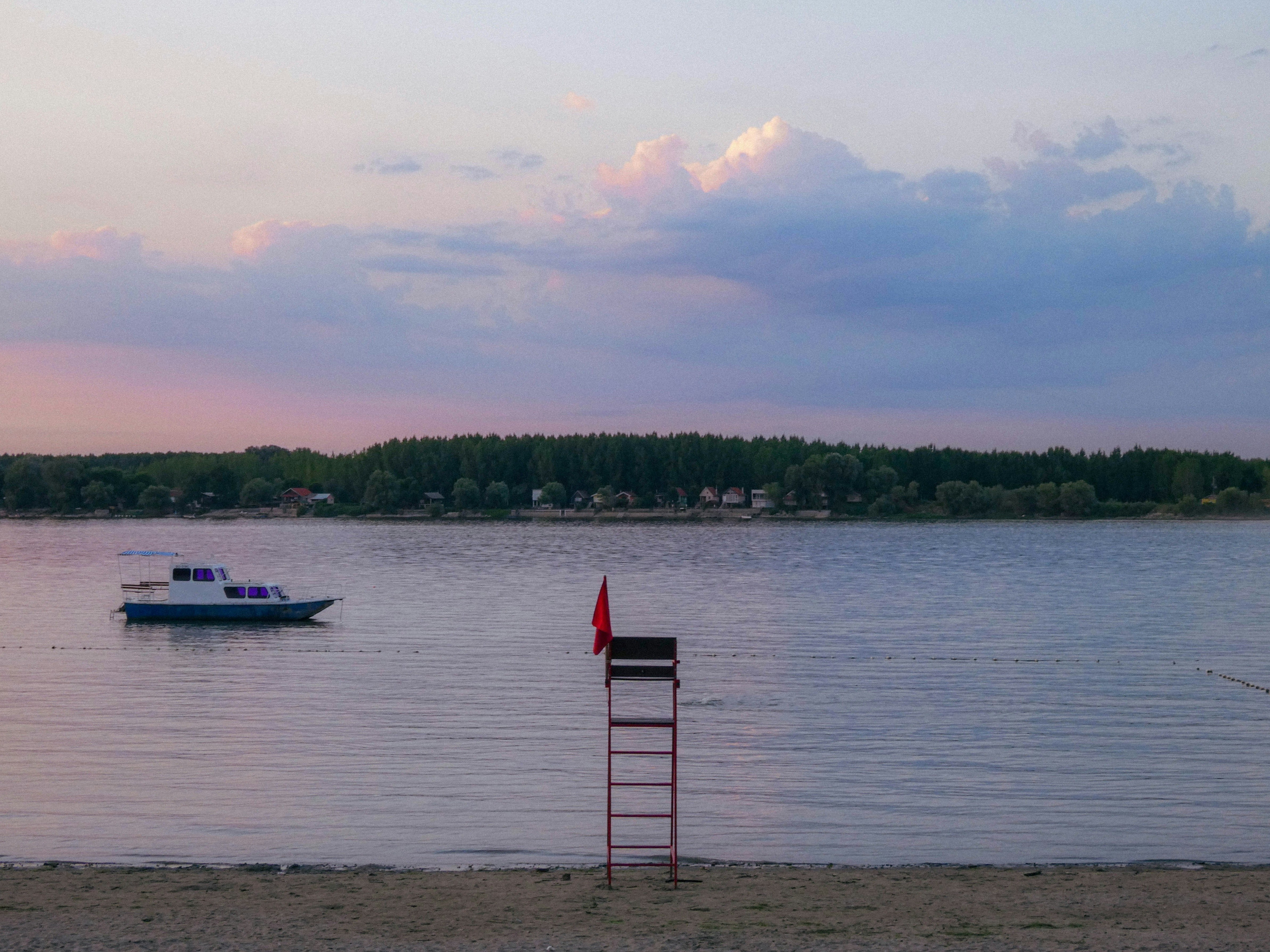 A calm evening on the beach, with a red lifeguard chair and a lone boat floating on the Danube under soft purple-blue clouds | Lifeguard chair on beach with boat on water