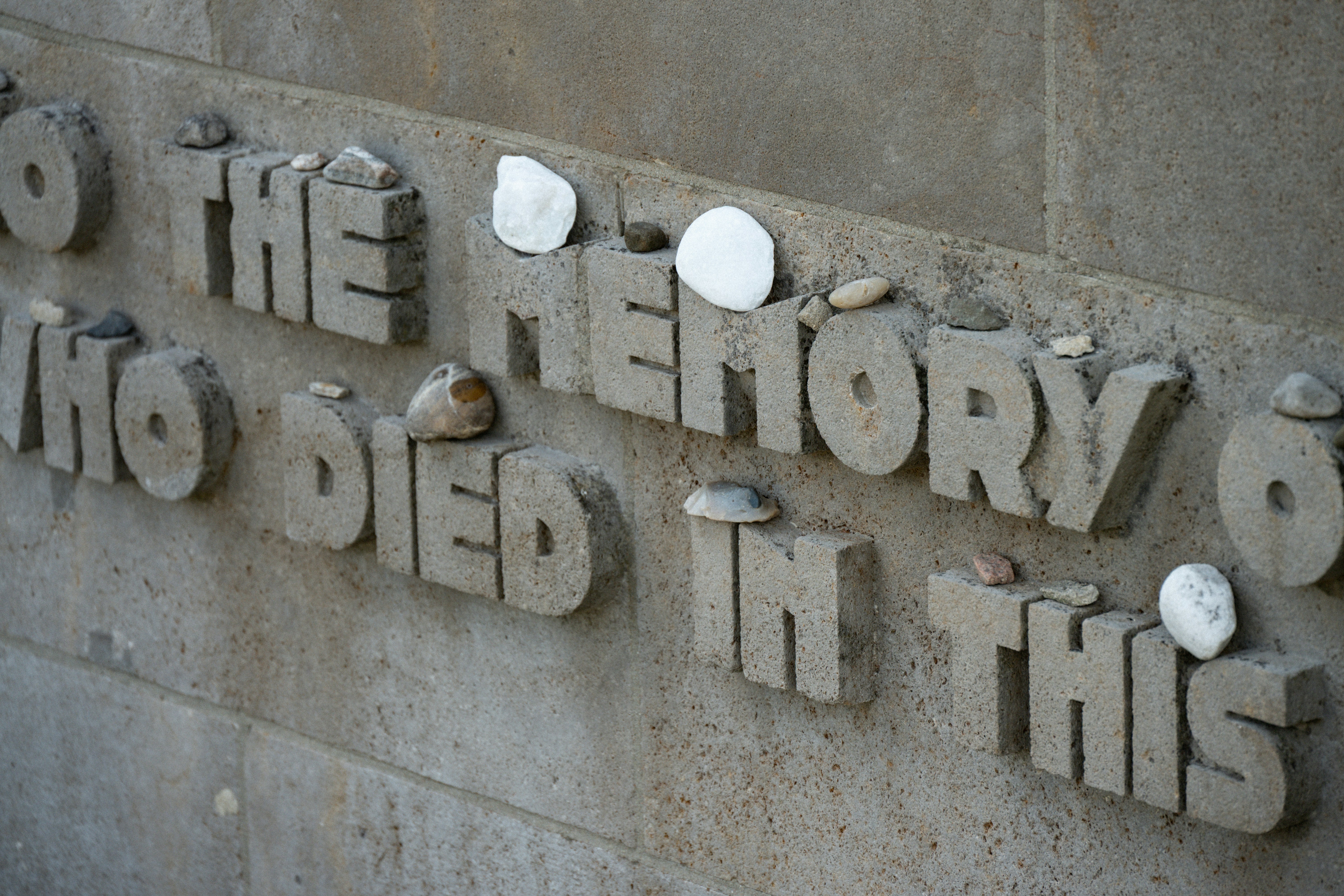 Concrete memorial inscription adorned with small stones, honoring those who perished. The arrangement of stones adds a personal touch to the tribute.