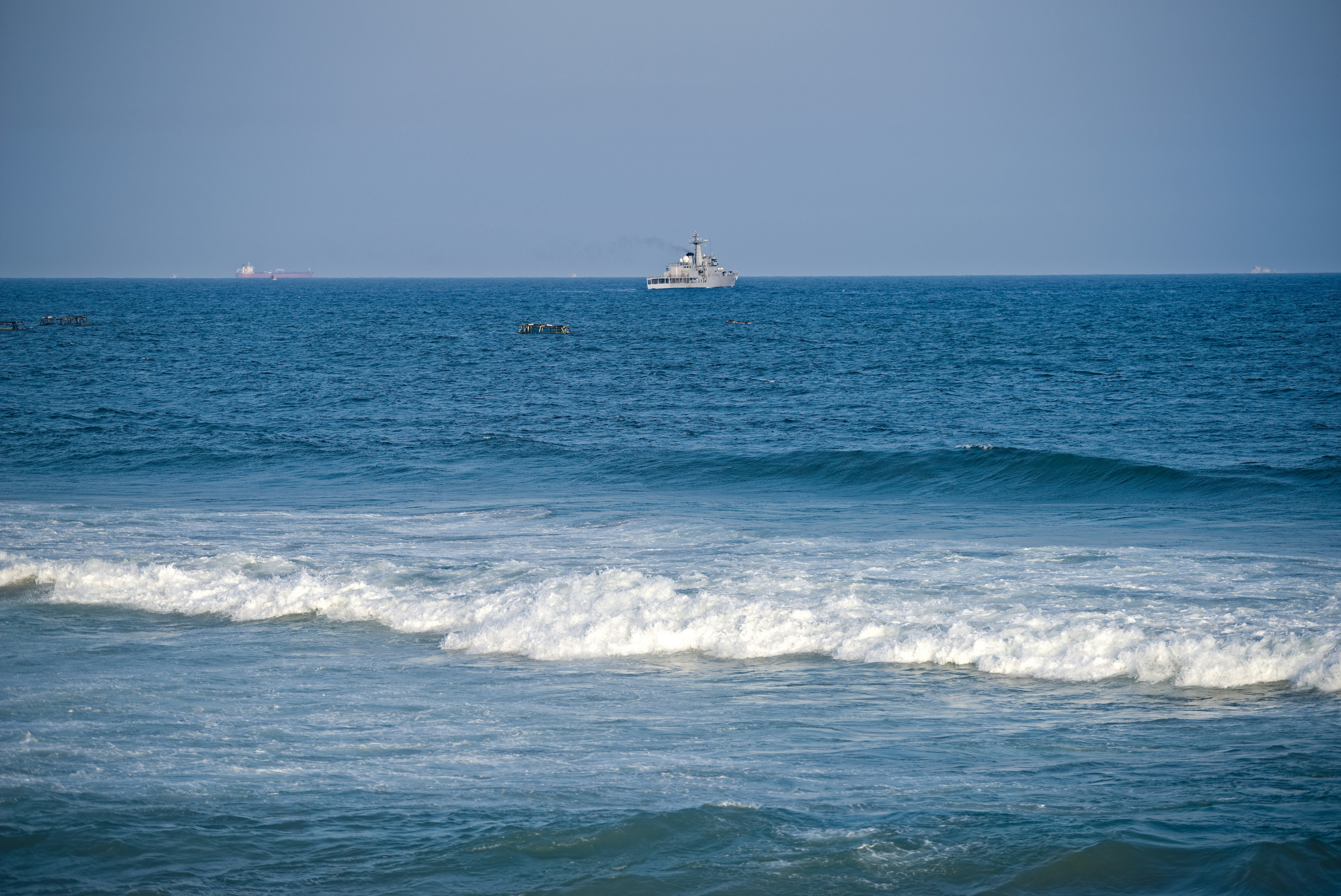 A distant ship navigates the calm ocean waters, with gentle waves rolling onto the shore under a clear blue sky.
