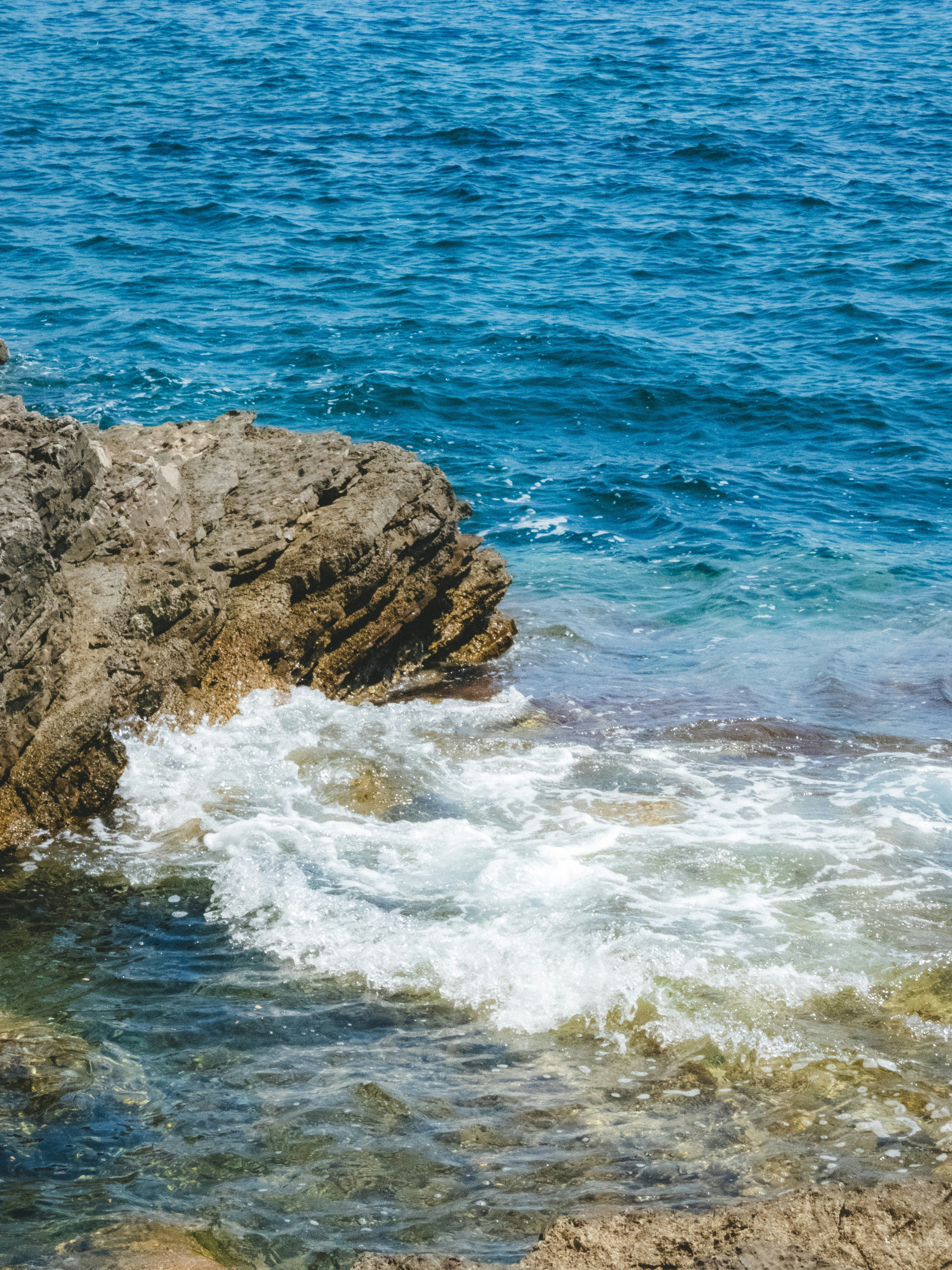 Clear turquoise water splashing against rugged coastal rocks | Waves crash against rocky shore under blue sky