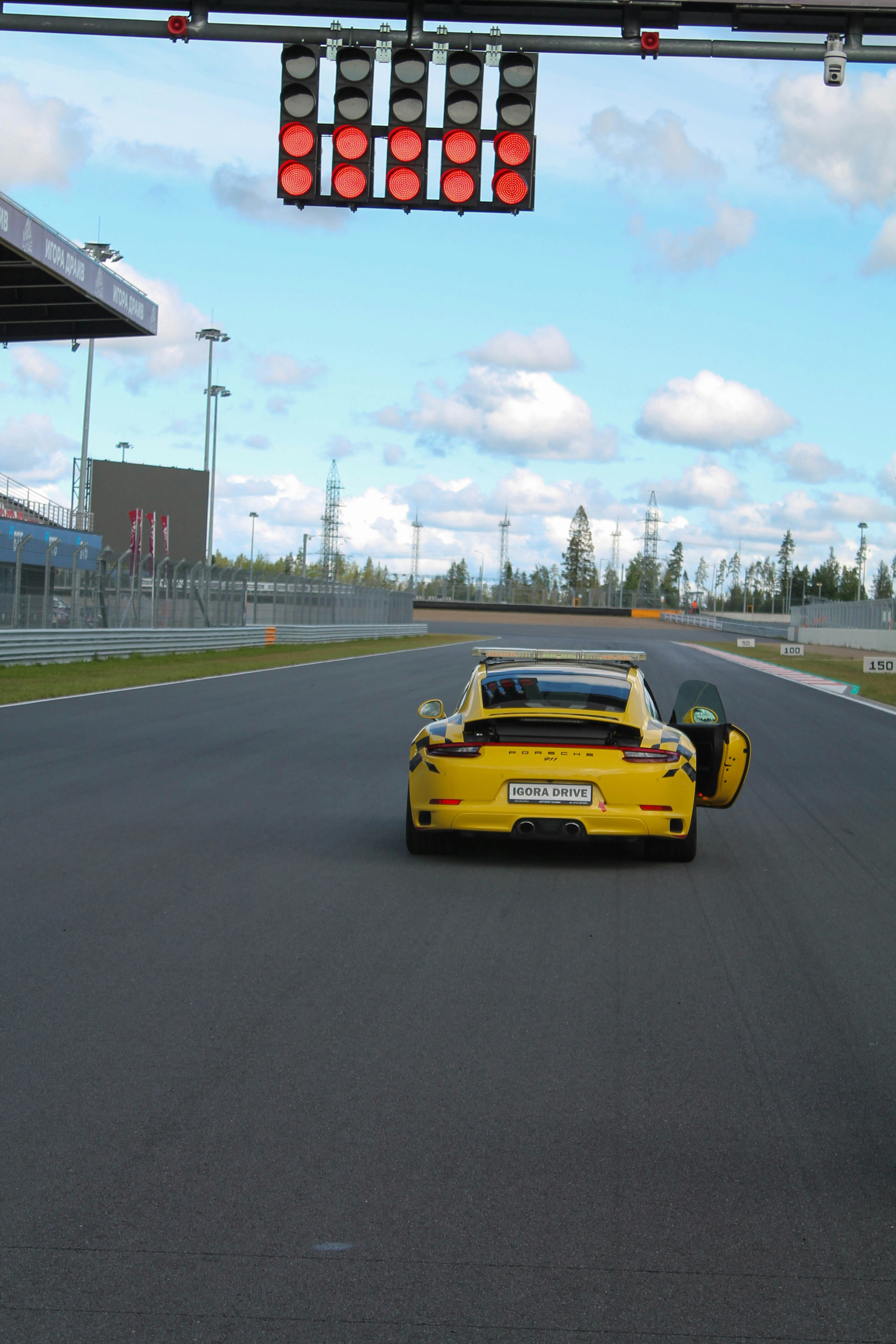 A vibrant yellow sports car prepares for a race on a track, with its door ajar and traffic lights signaling the start. The backdrop features a clear sky and distant trees.