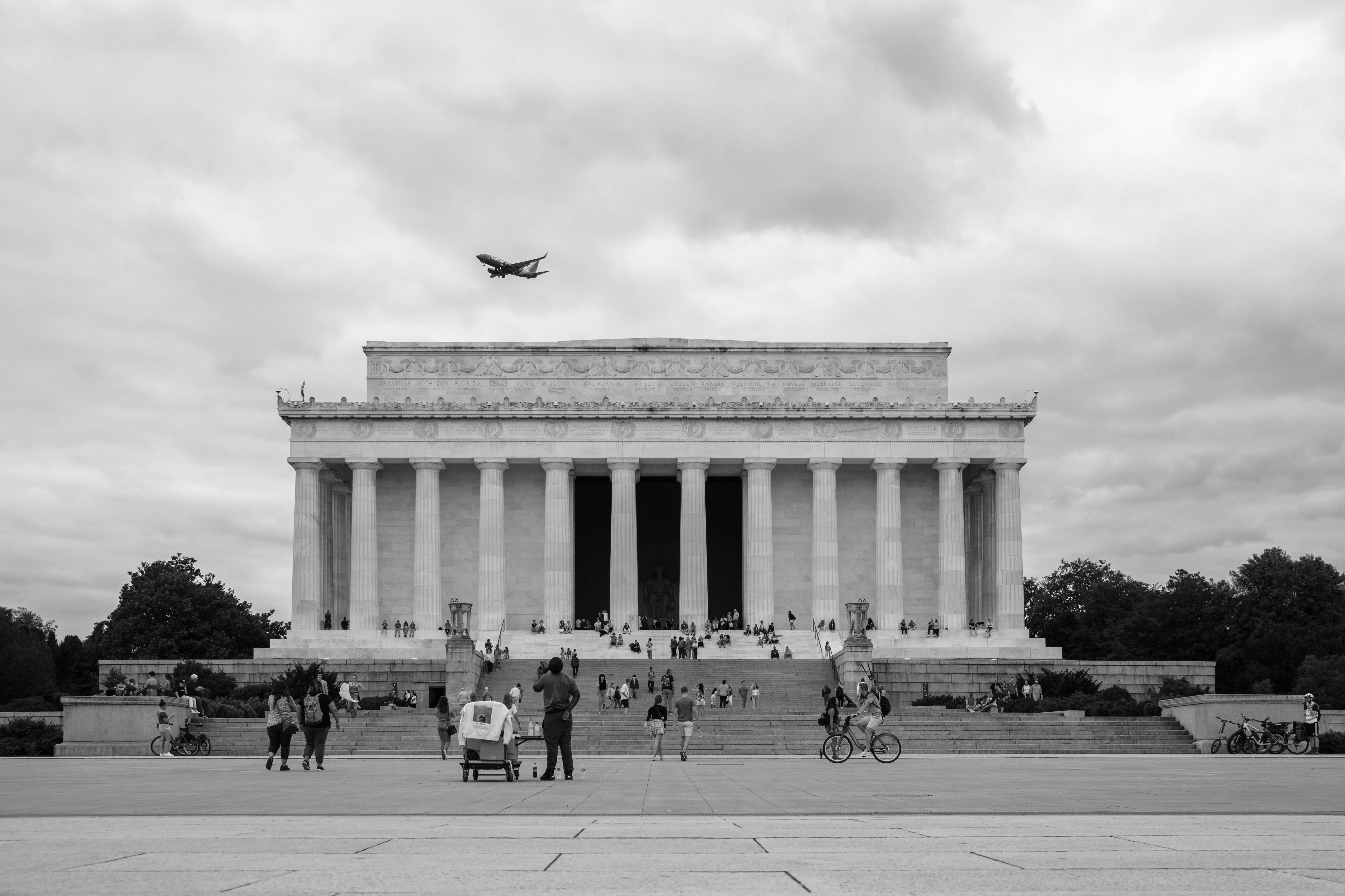 Visitors are scattered around the Lincoln Memorial steps and plaza in Washington, DC. A commercial plane flies overhead on a cloudy day. | Lincoln memorial with people and airplane overhead