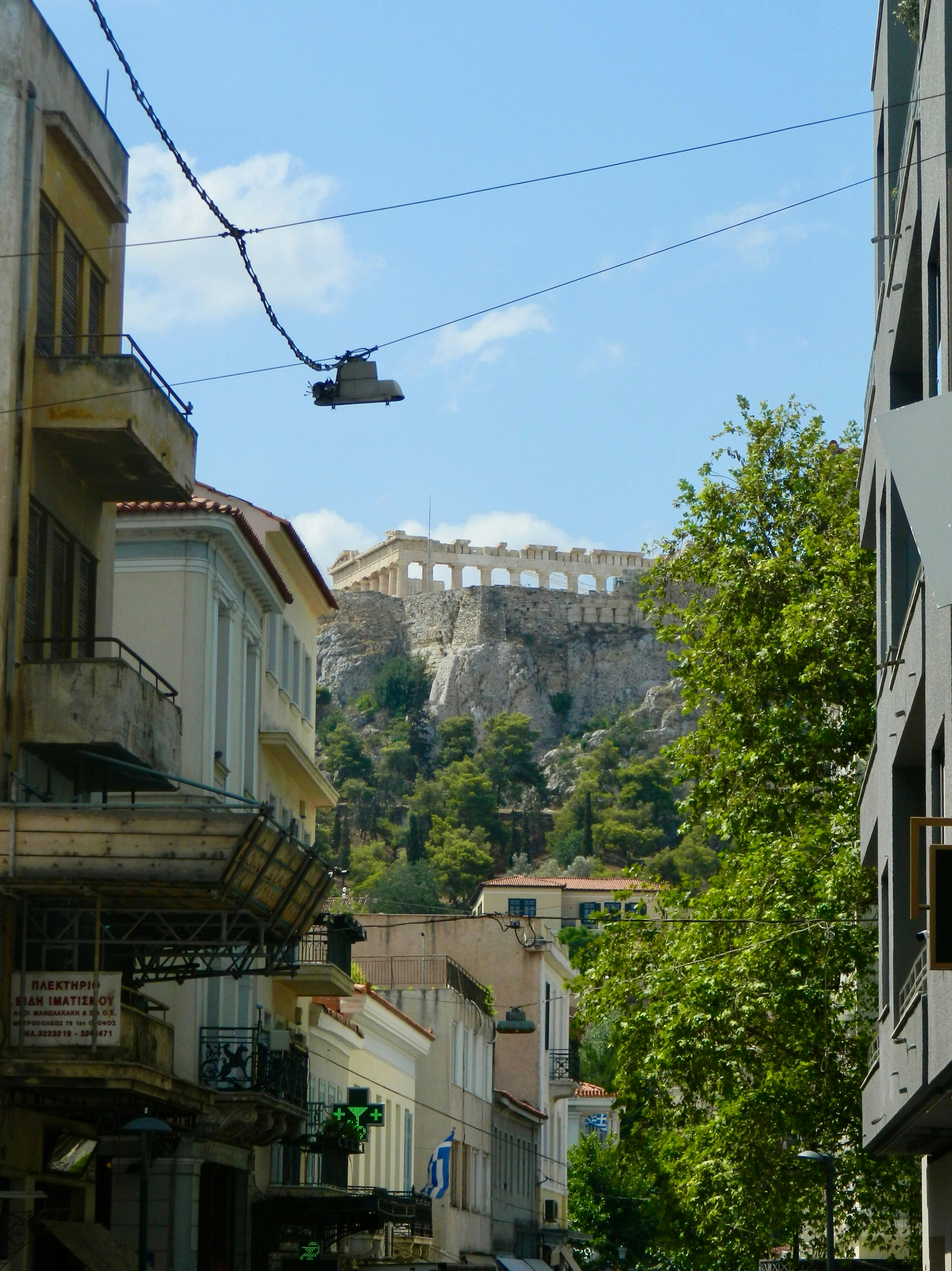 The Acropolis stands majestically above a bustling street, framed by modern buildings and lush greenery.