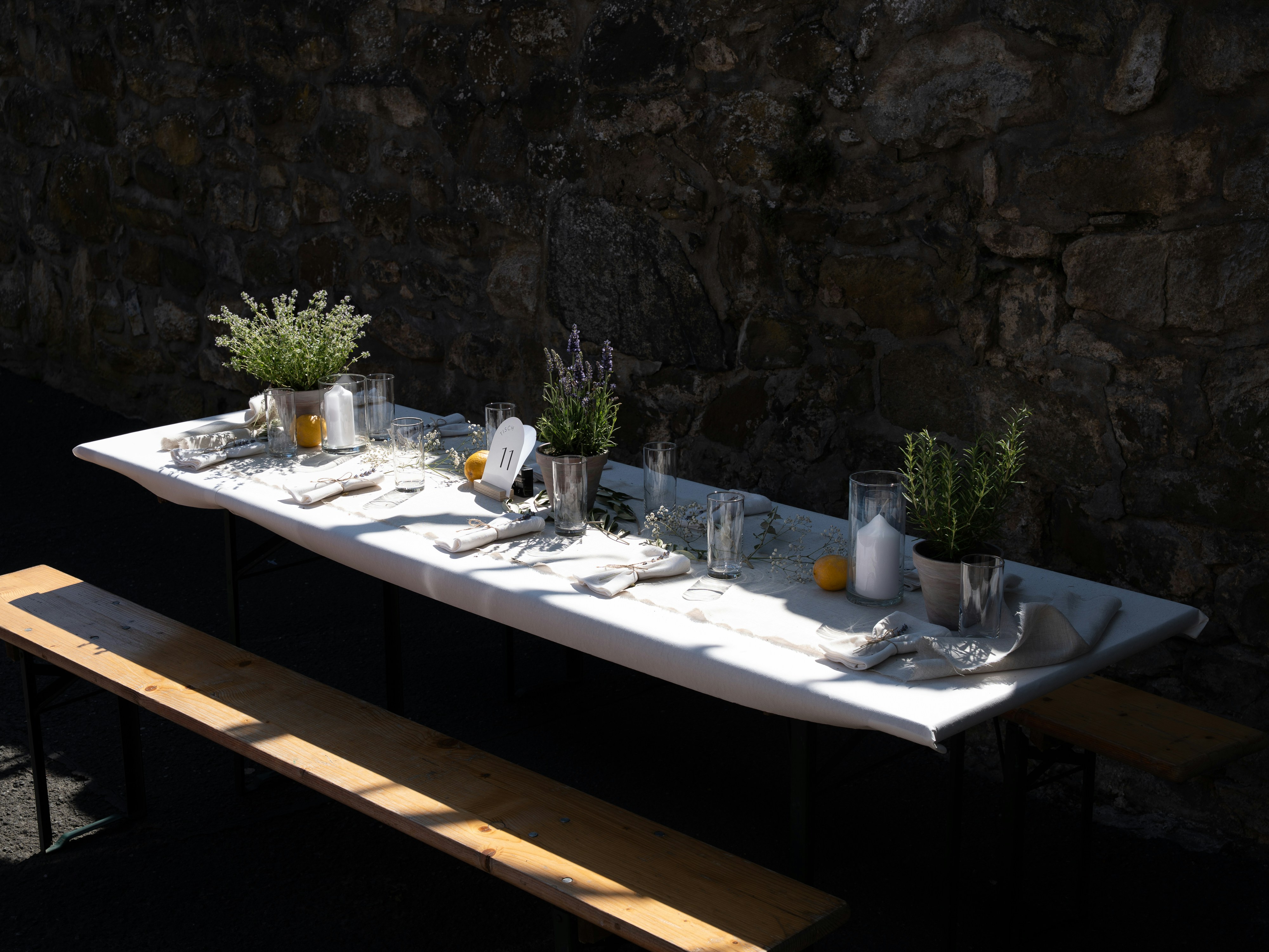 Long dining table set with glassware, napkins, and potted herbs against a rustic stone wall. A lemon adds a touch of color.