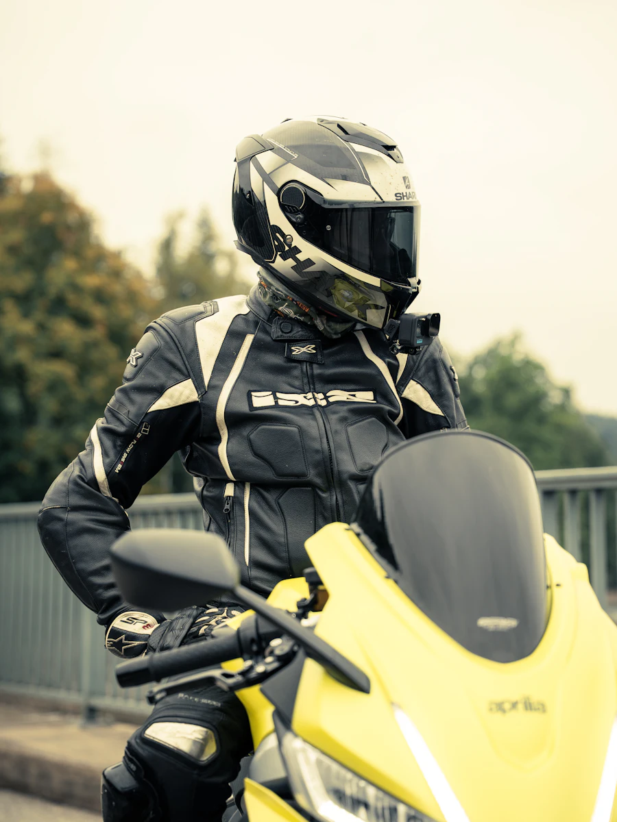 Helmeted motorcyclist in black leather jacket standing over a yellow sportbike with visor down and an action camera mounted.