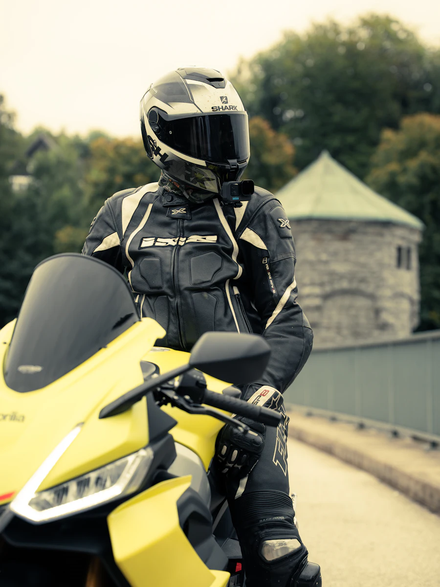 Helmeted motorcyclist in black leather gear seated on a bright yellow sportbike with a stone tower and leafy background.