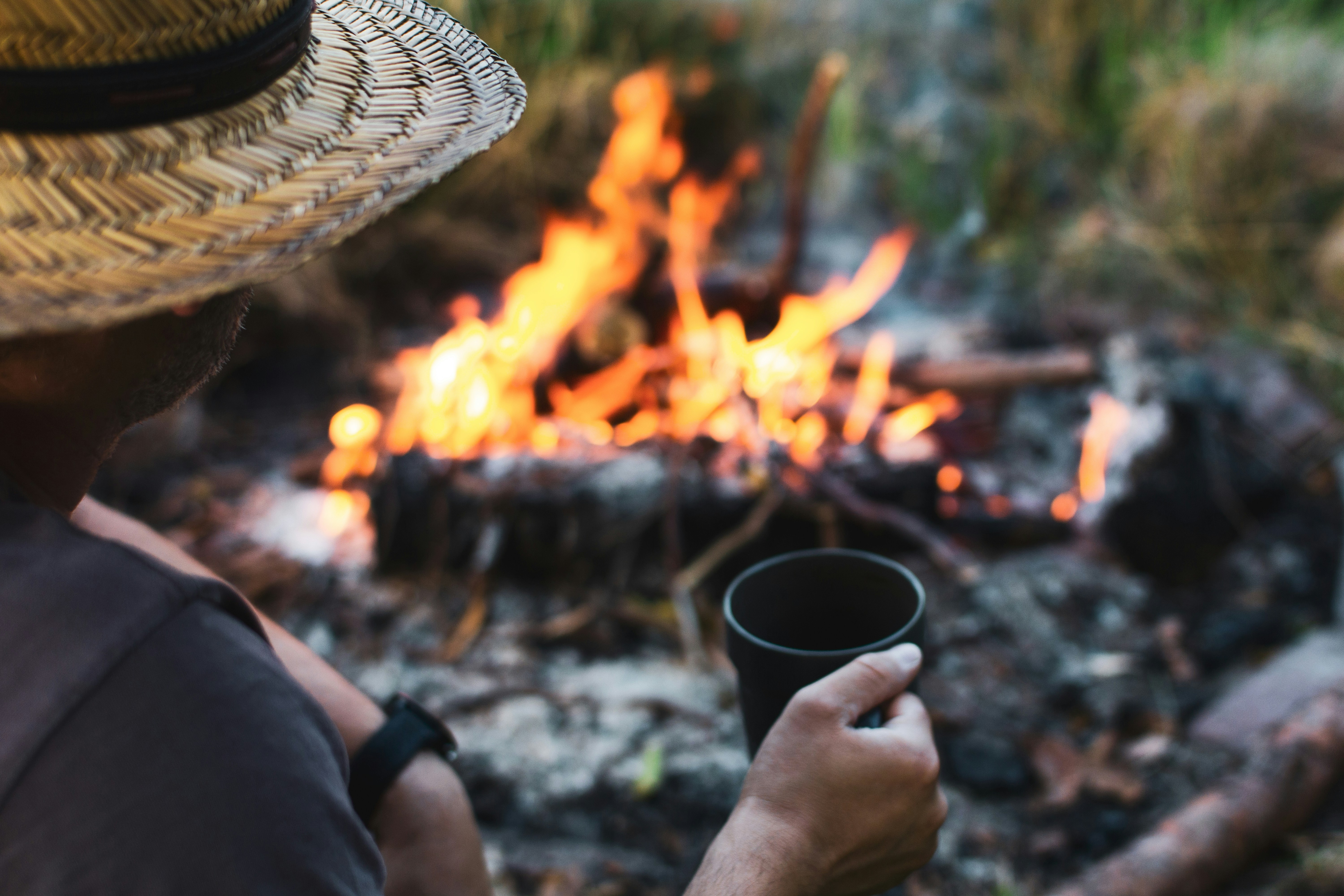 Camping, outdoor | Person in hat holds mug near campfire outdoors