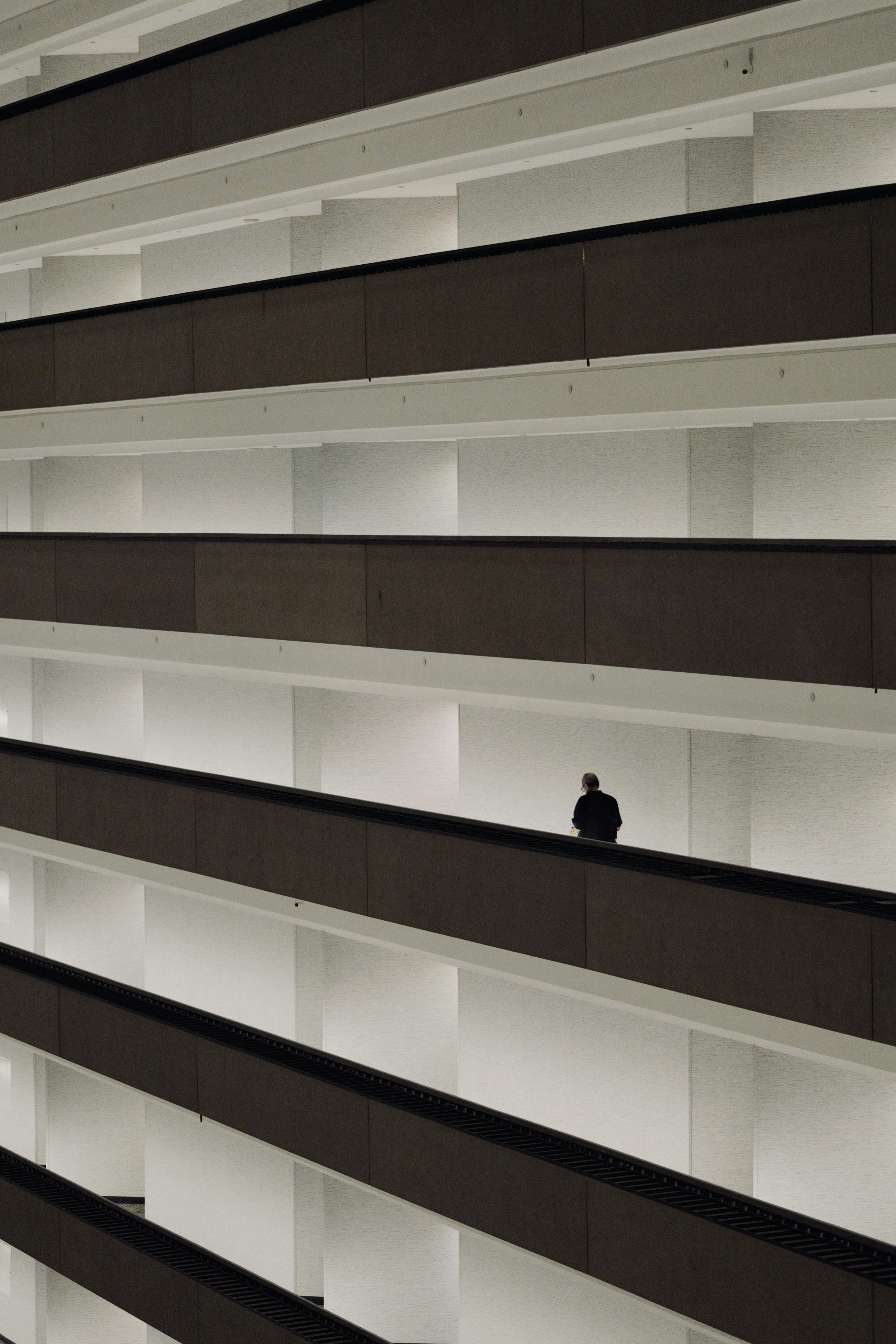 A lone figure stands on a balcony, framed by the stark, geometric lines of a modern building's interior. The soft illumination enhances the sense of solitude.