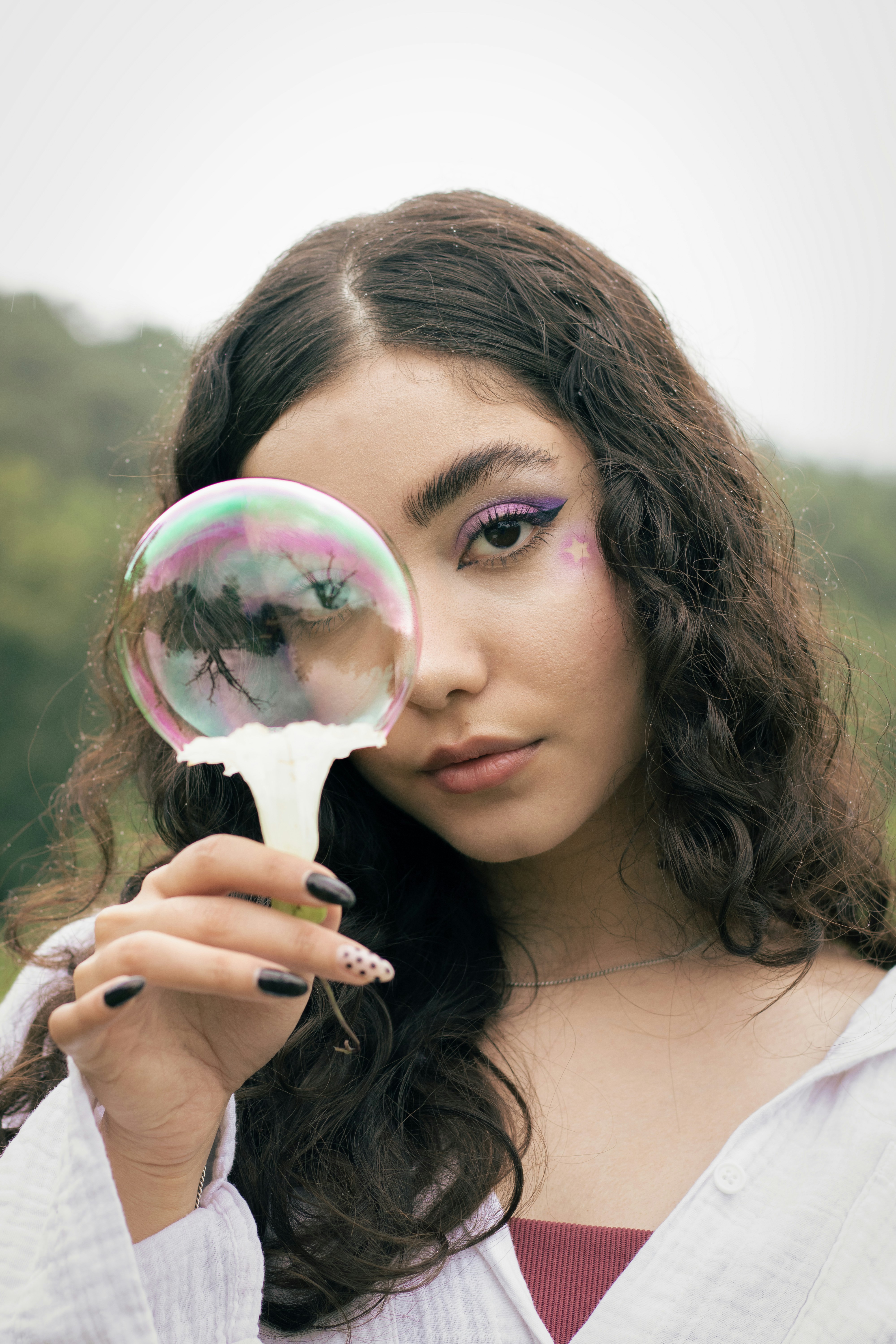 Young woman gazing through a bubble, showcasing intricate reflections and vibrant colors against a blurred natural backdrop.