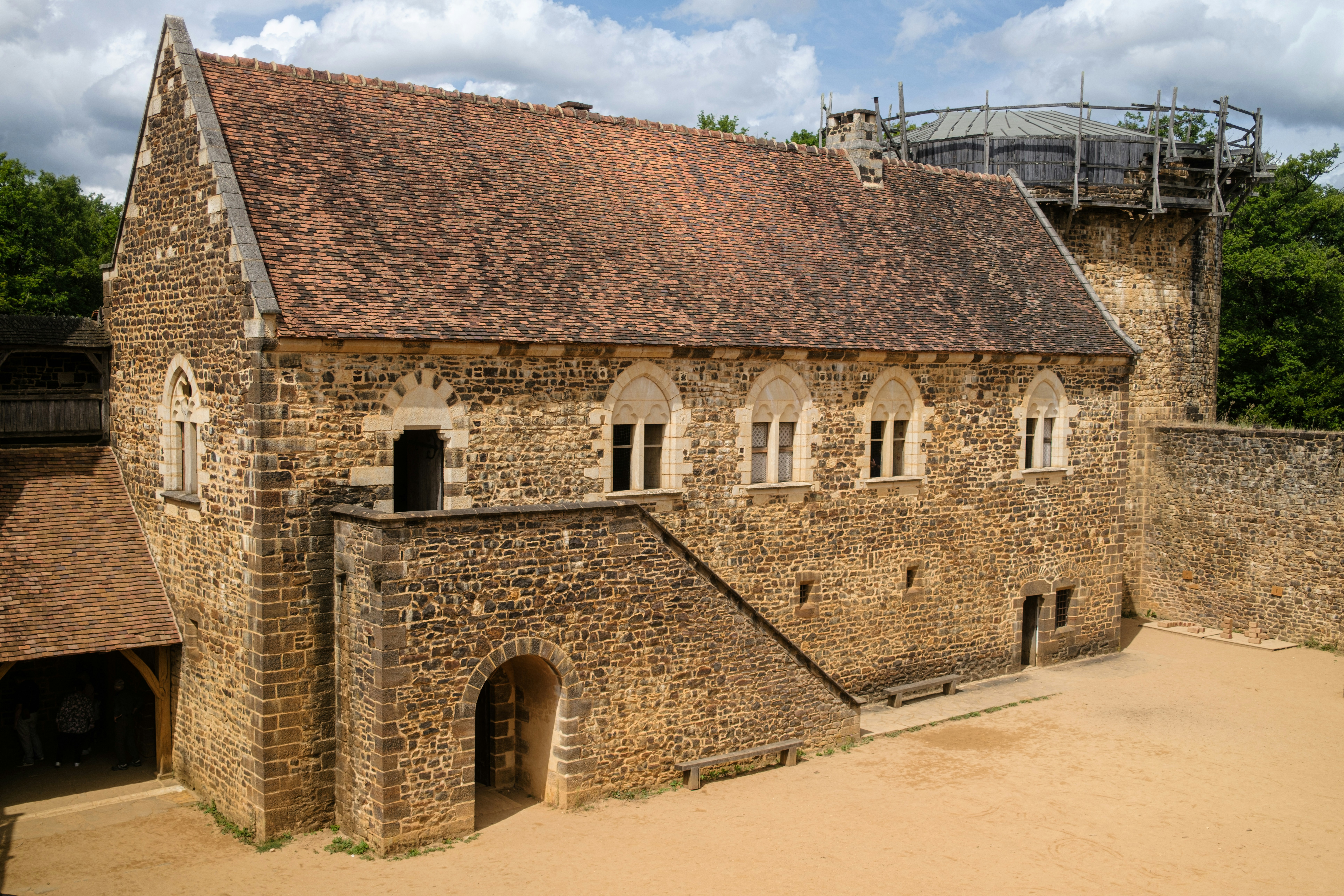 Medieval stone building with arched windows and a weathered roof, showcasing historical architecture. A partially constructed tower looms in the background.