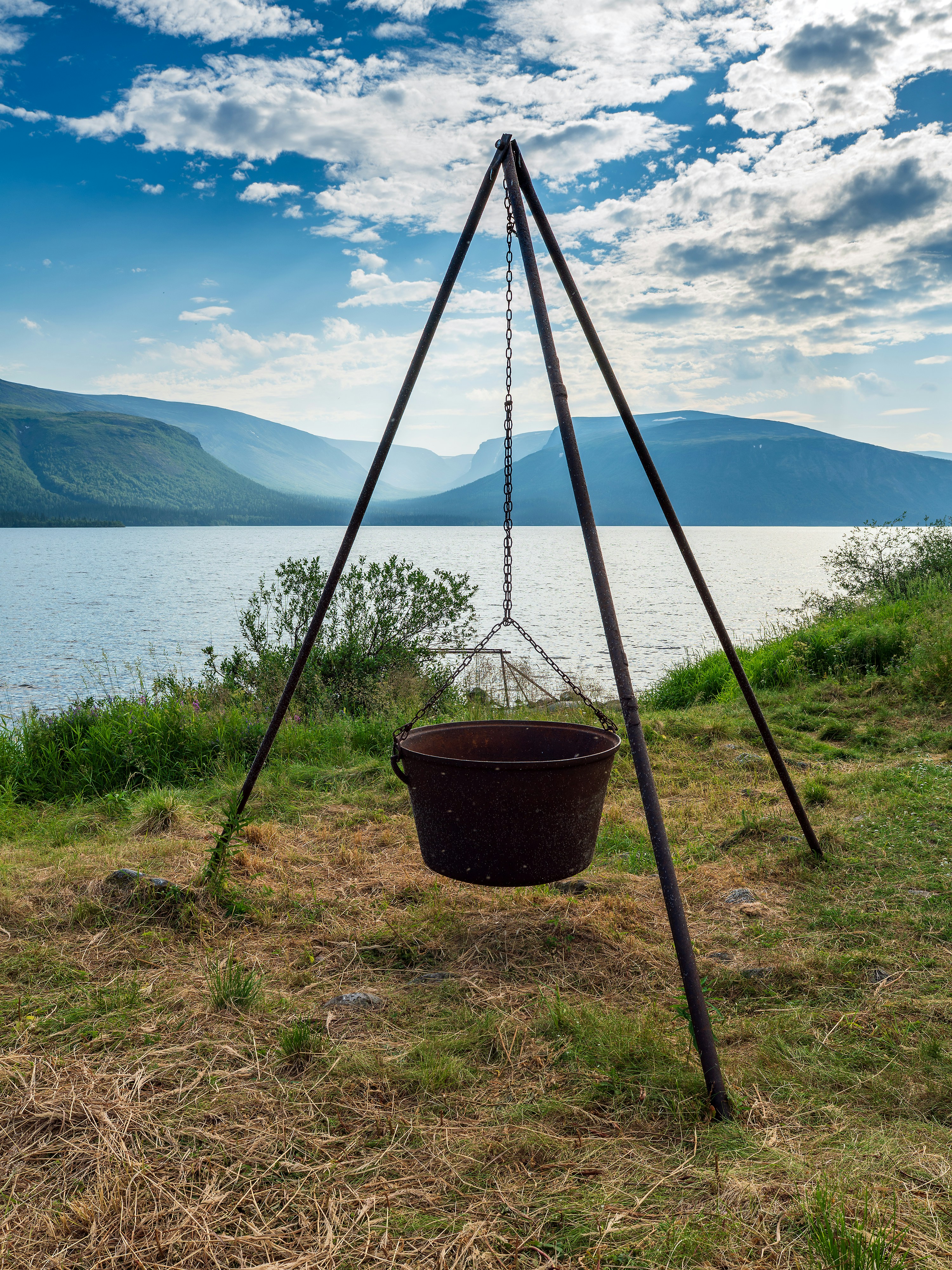 Tripod with cauldron over grassy landscape and lake.