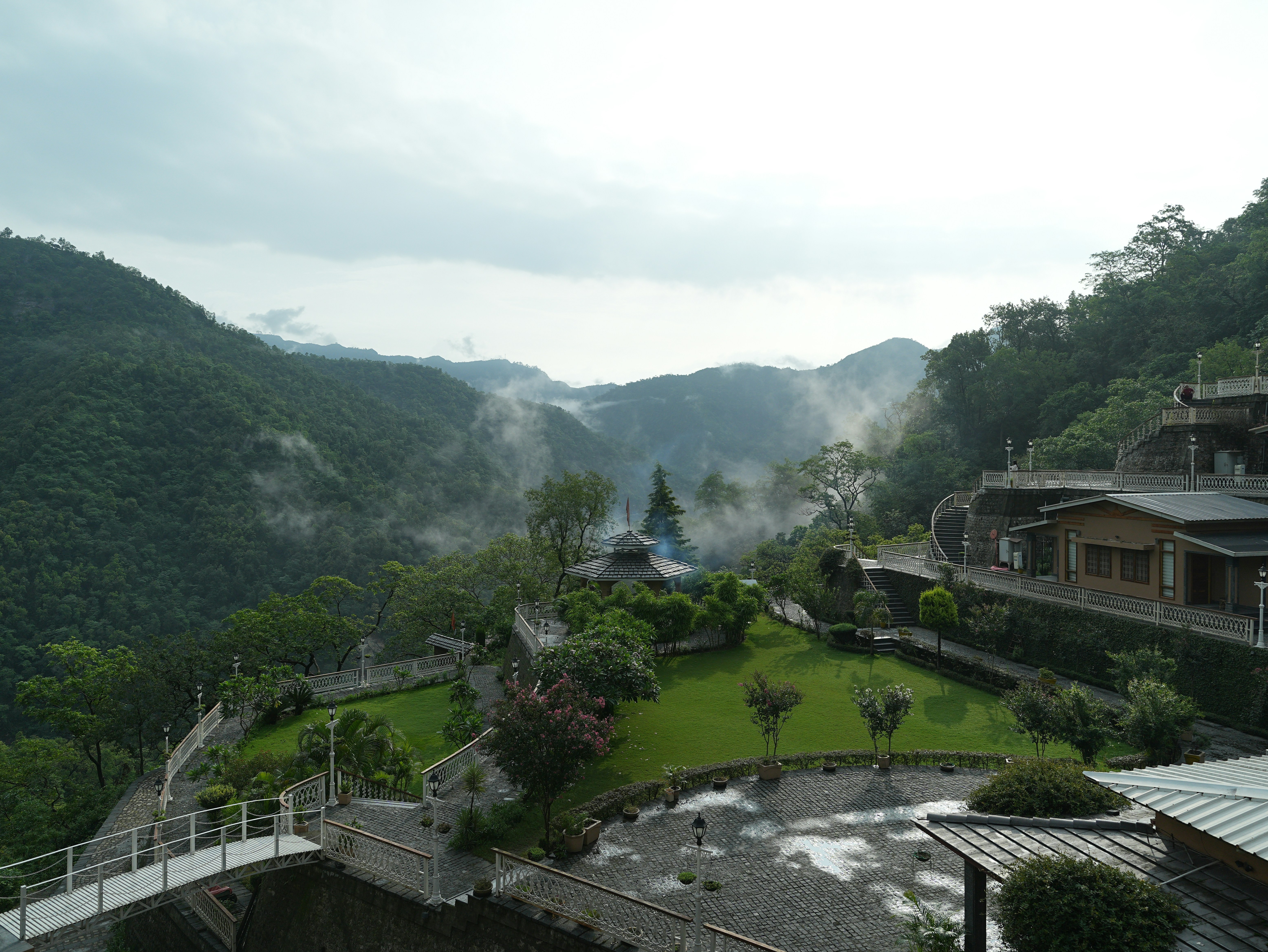 Lush green landscape with a well-manicured garden and modern structures, set against a backdrop of misty mountains.