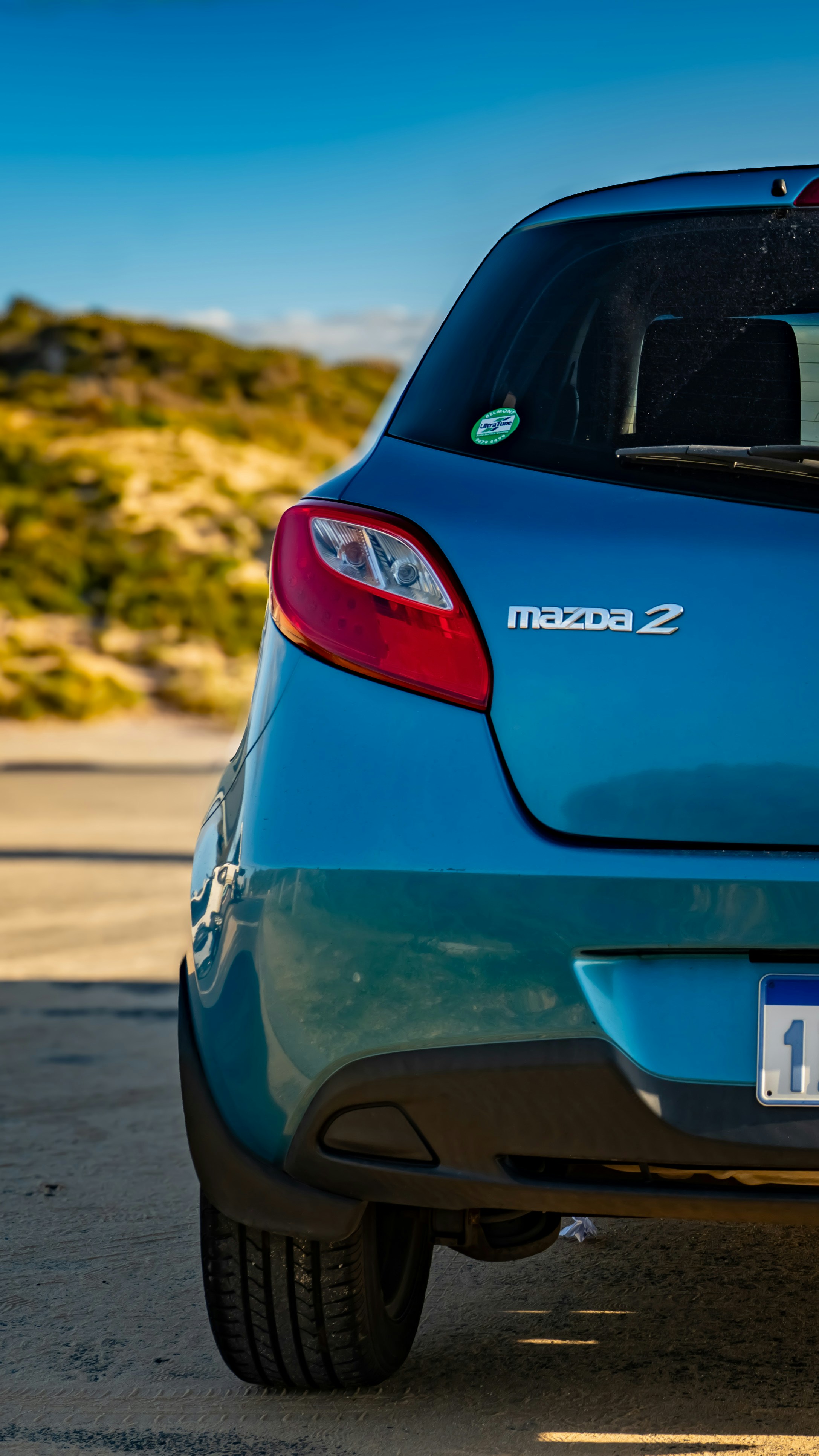 Rear view of a Mazda 2 parked on a sandy surface, showcasing its sleek lines and vibrant blue color. The background features a natural landscape.