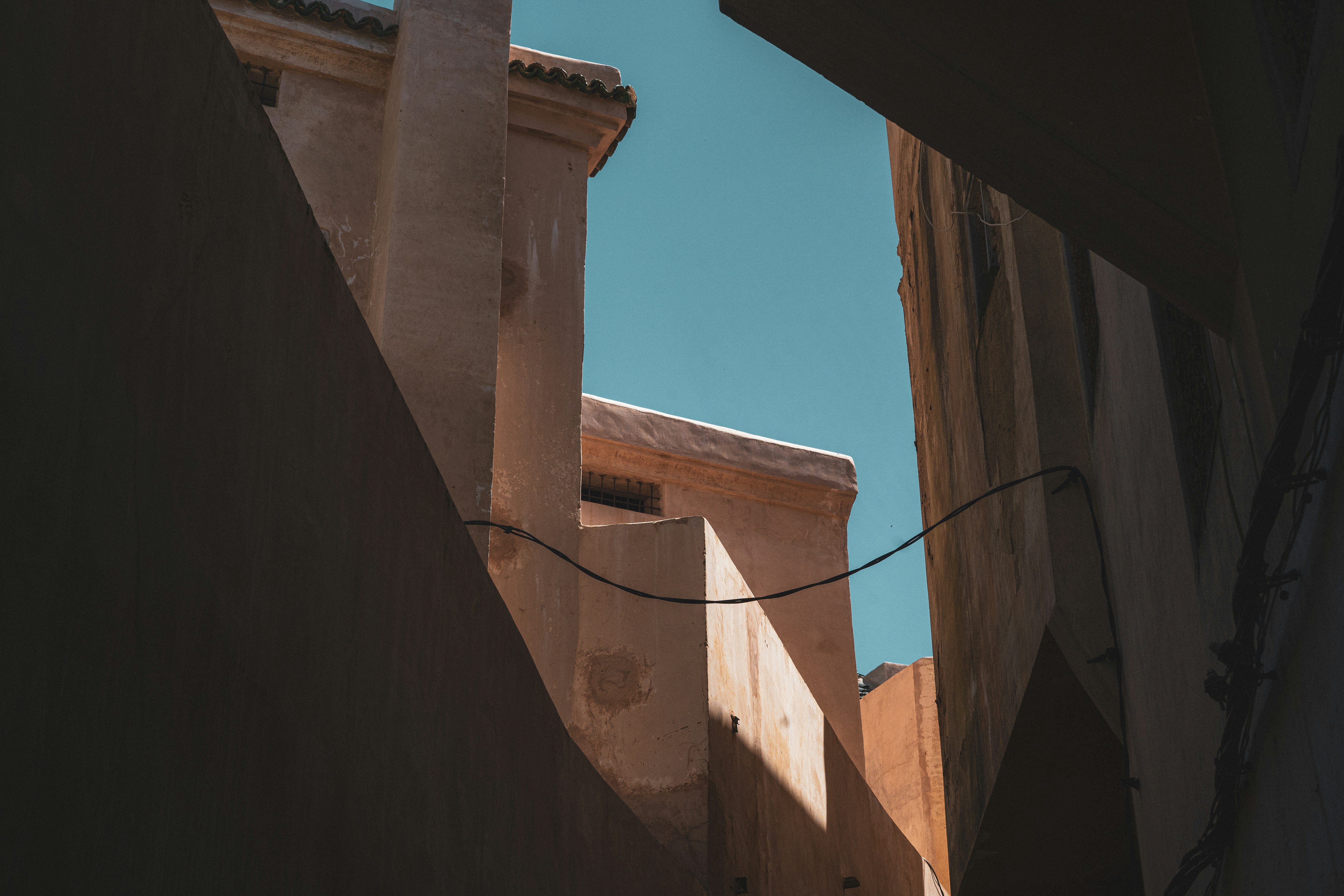 Narrow alleyway revealing a glimpse of blue sky framed by warm, textured walls. The interplay of light and shadow creates a dynamic urban scene.
