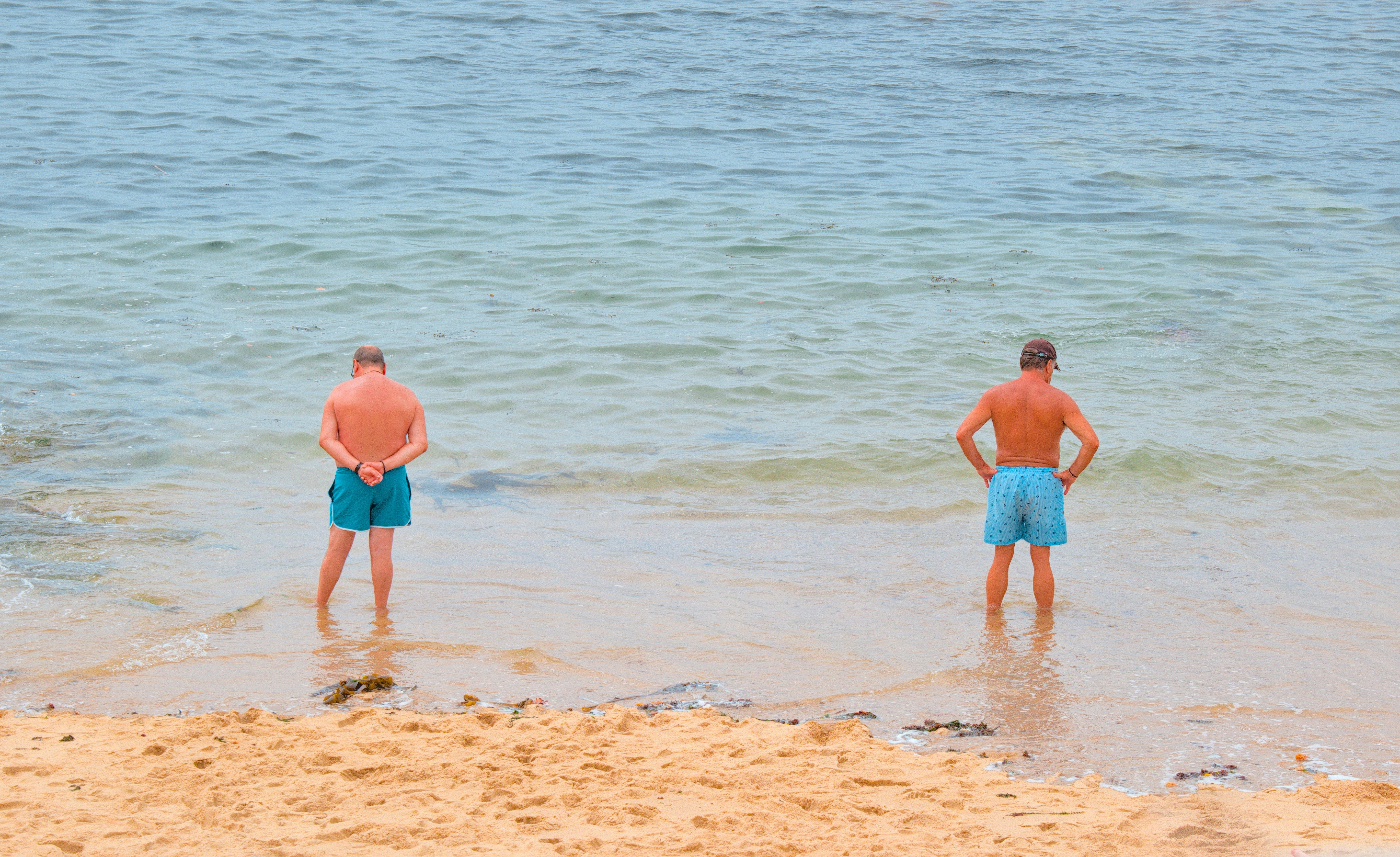 Two men in swim trunks stand on a sandy beach.