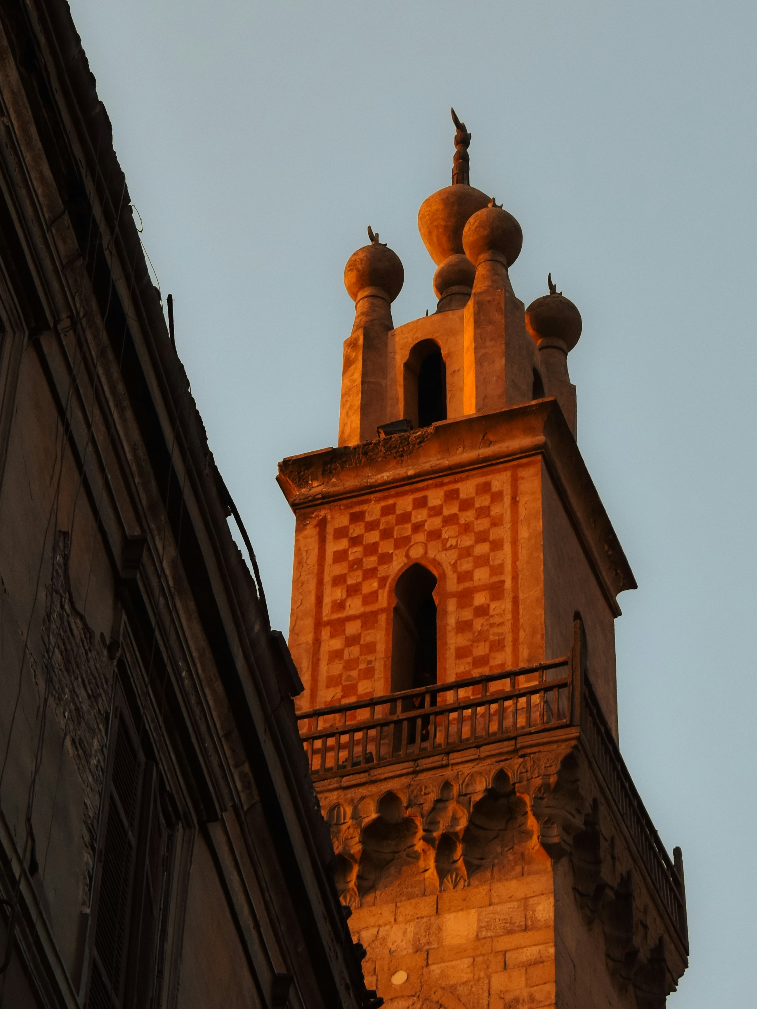 A historic tower bathed in warm, golden light as the sun sets, highlighting intricate brickwork and a stepped, checkerboard pattern. The sunlit facade contrasts with the cool sky, emphasizing the architectural details and vertical silhouette of the tower.