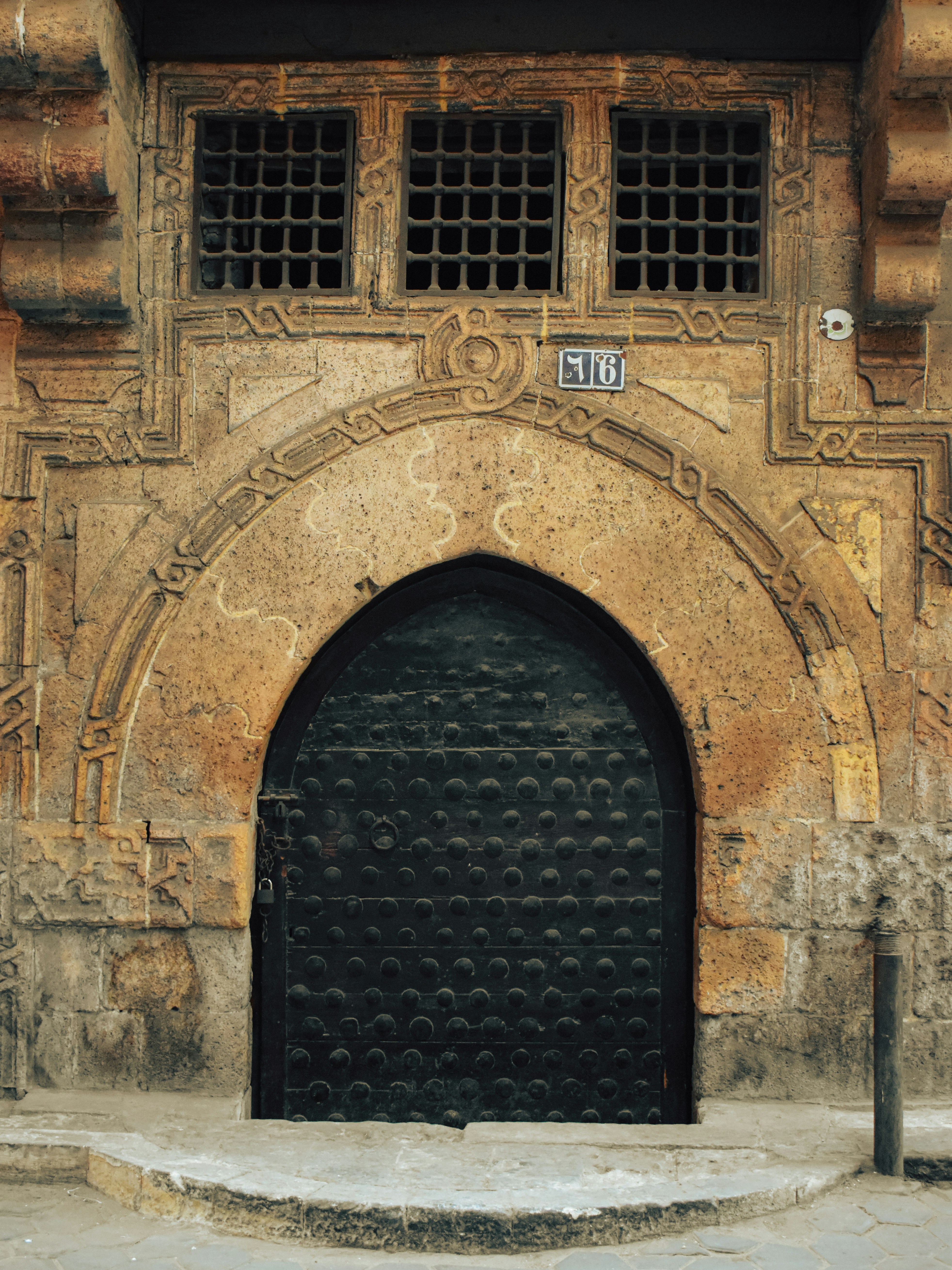 A weathered, arched doorway set in a sun-baked stone façade. The heavy black door is studded with round metal rivets, flanked by intricate geometric carvings and an ancient, worn frame. Small lattice windows above add a hint of mystery, while the surrounding stone shows rich textures and warm earth tones. This image captures historic architectural detail, craftsmanship, and a sense of timeless fortification. Ideal for themes of history, heritage, architecture, and vintage or rustic aesthetics. | Ornate arched doorway with textured metal door