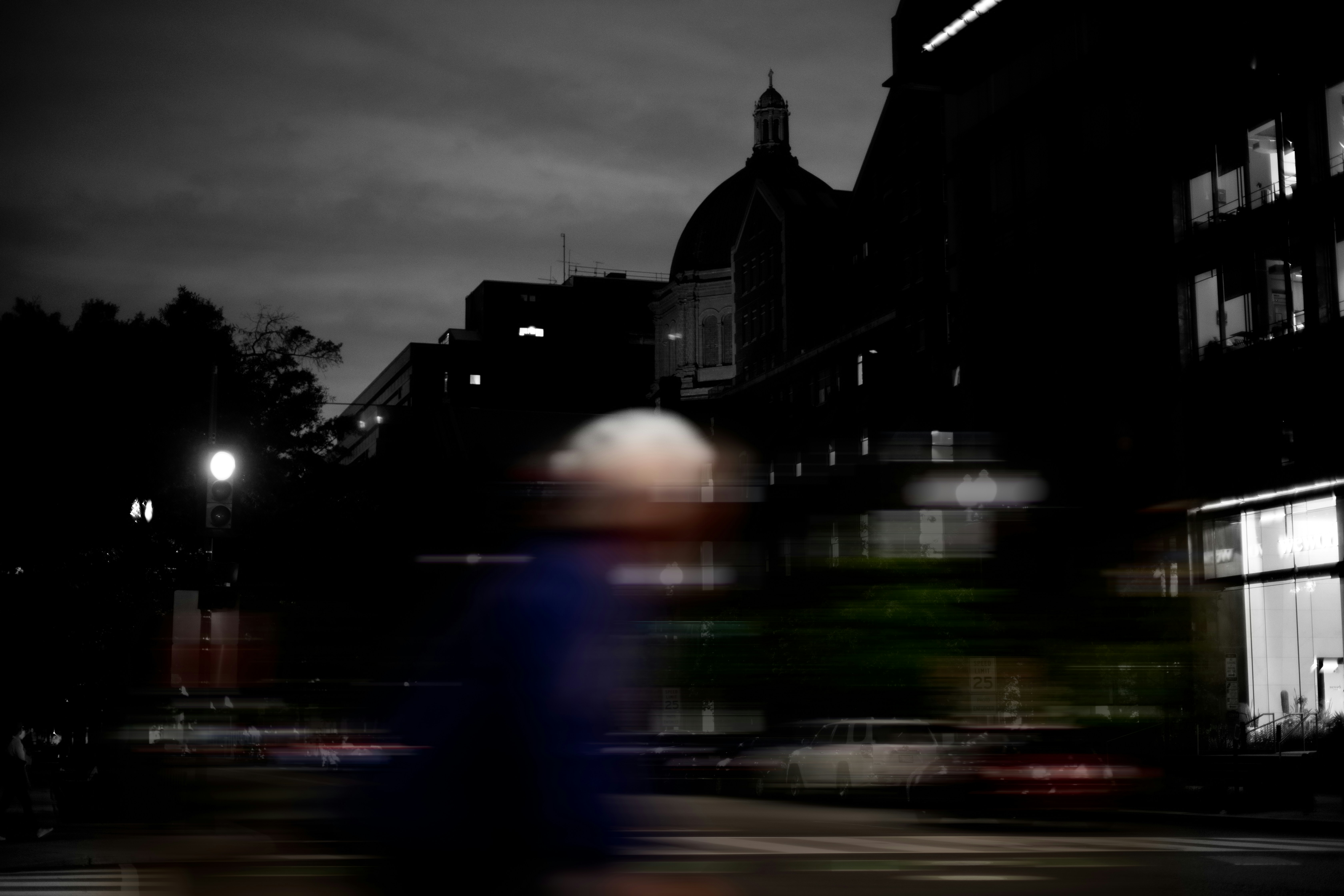 A blurred figure crosses a dimly lit street against a backdrop of urban architecture at dusk.