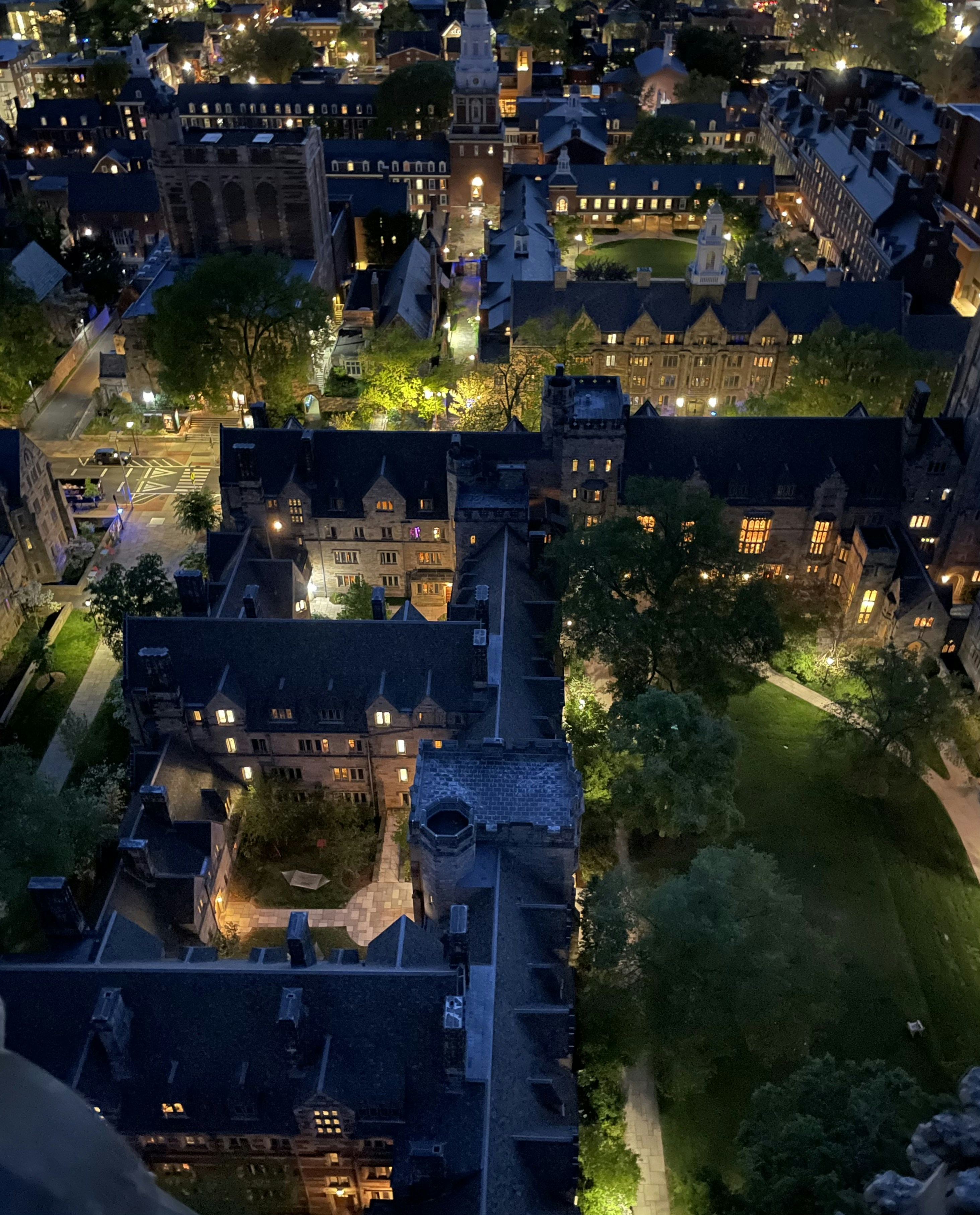 An aerial view of a historic campus illuminated at night, showcasing intricate architecture and lush greenery. The scene captures the interplay of light among the buildings and trees.