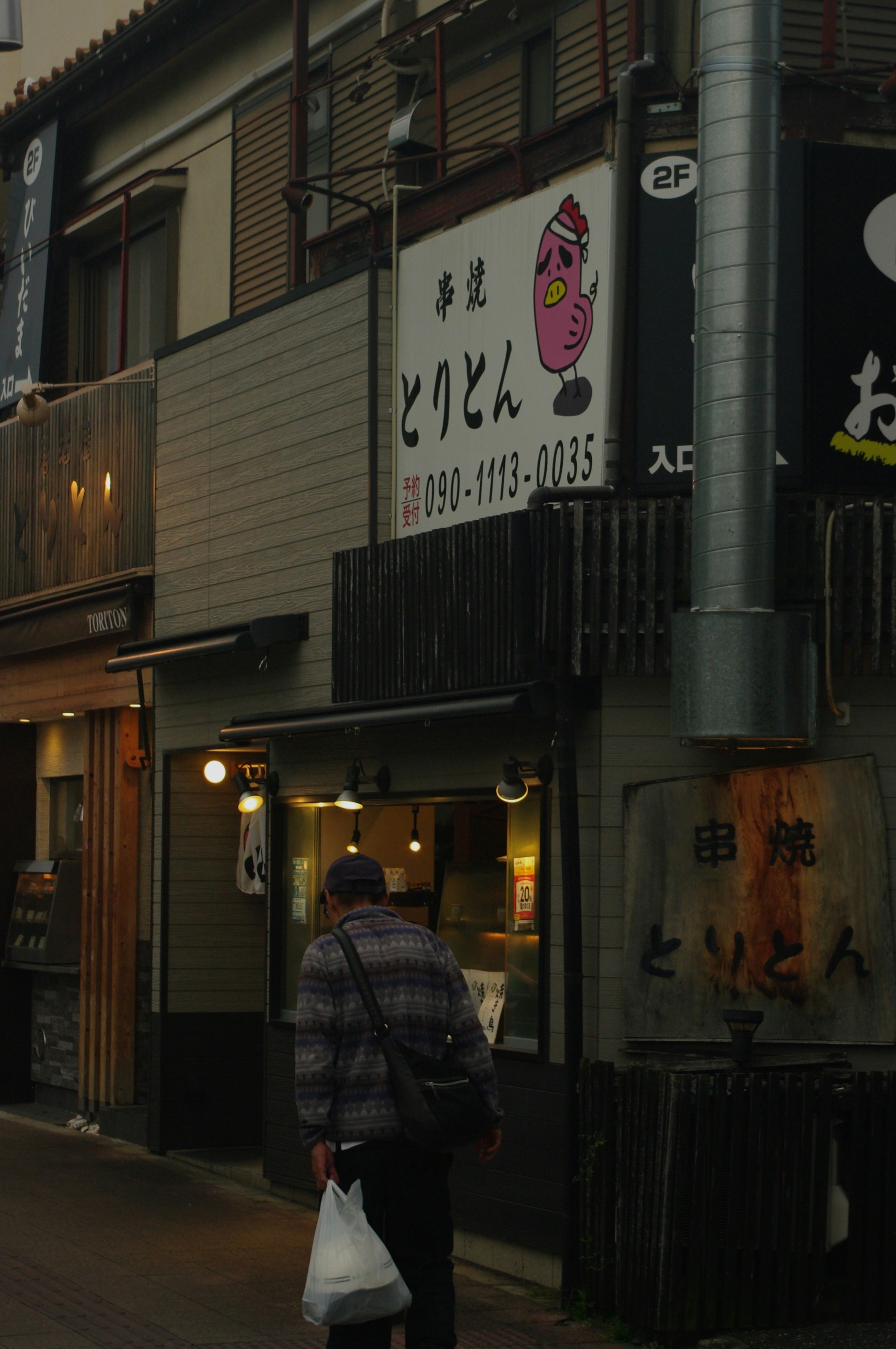 A man carrying a plastic bag walks past a yakitori shop with playful signage and warm lights. | Man walking past japanese restaurant with signage
