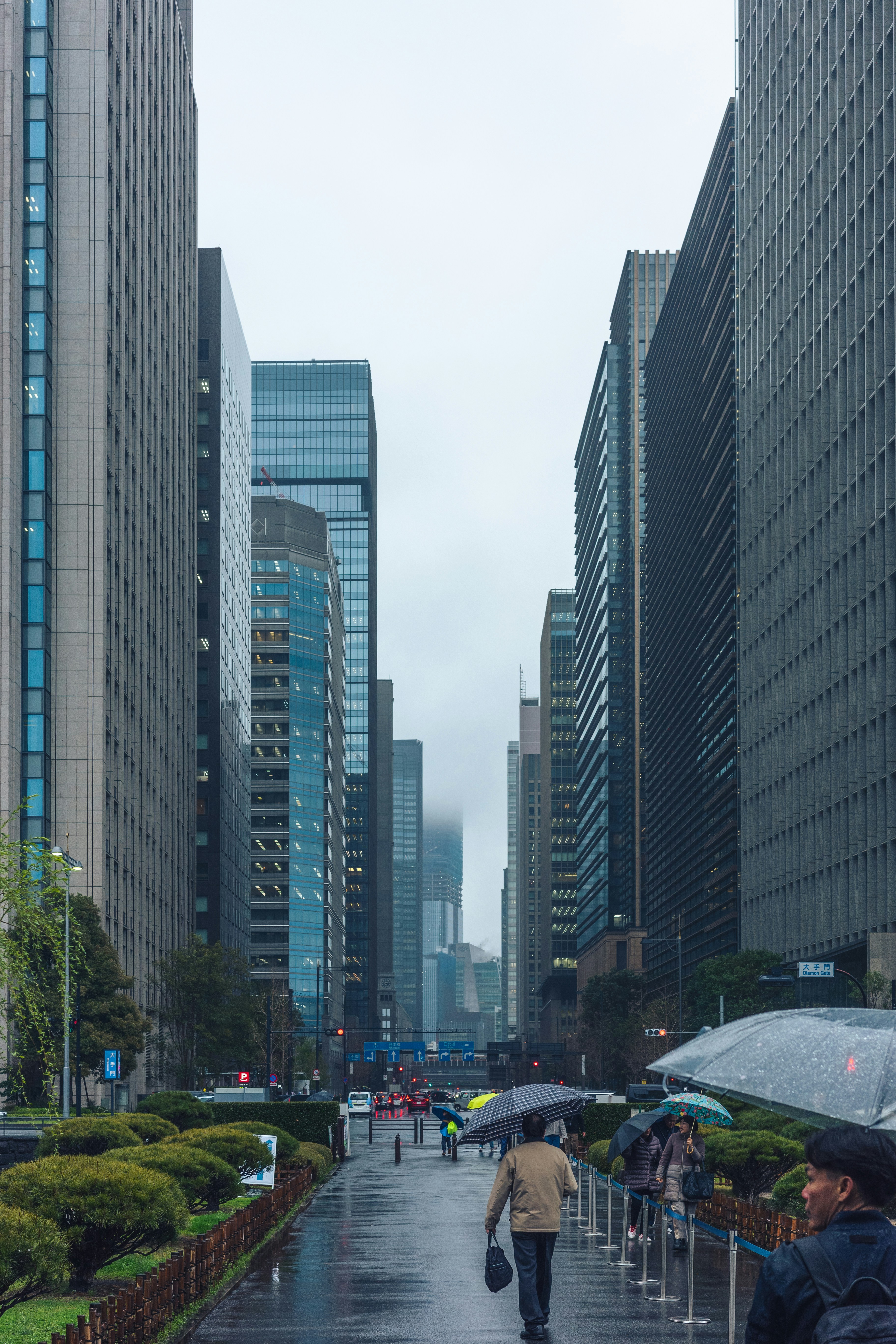 City street lined with towering skyscrapers under a cloudy sky, featuring pedestrians with umbrellas and reflections on wet pavement.