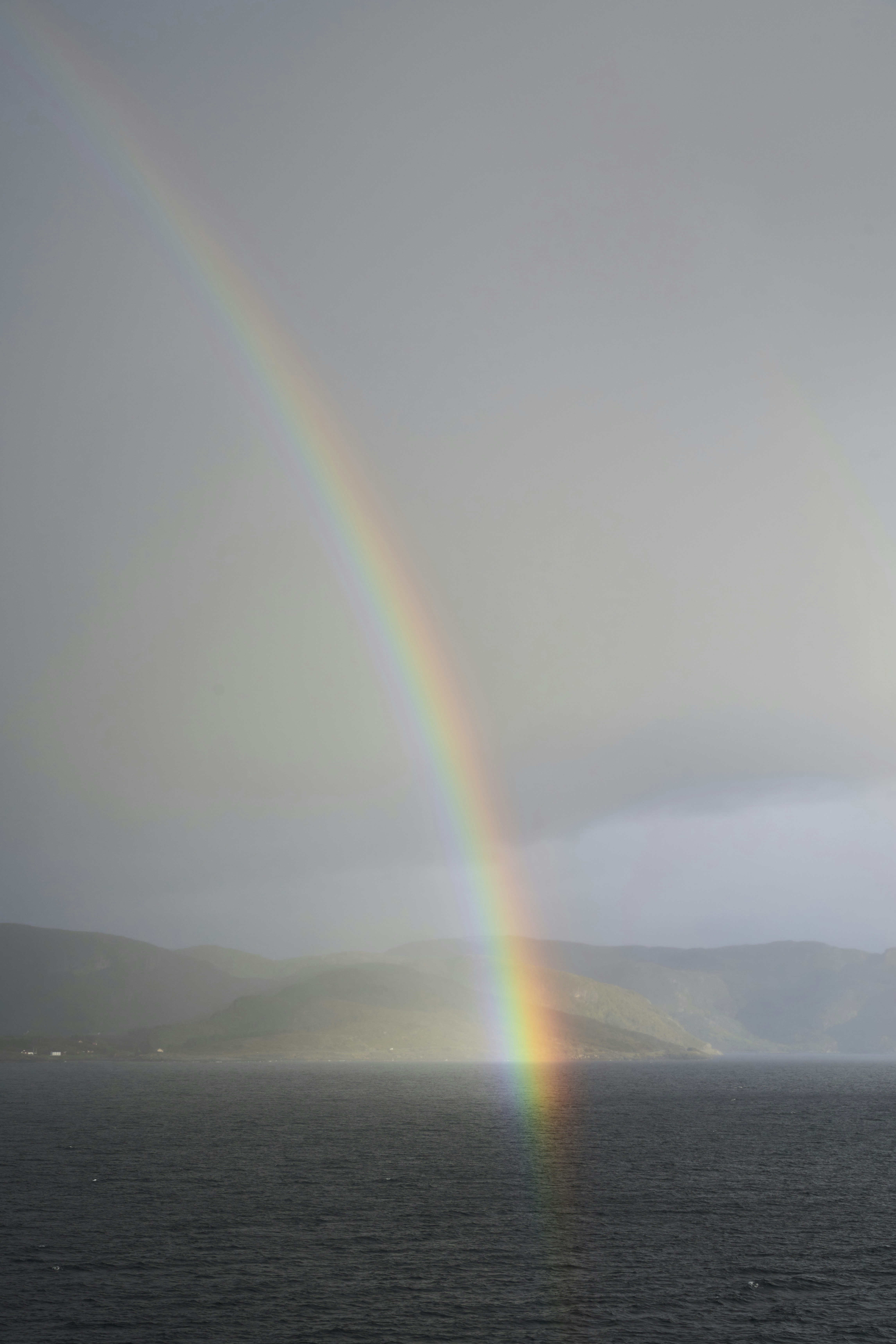 A vibrant rainbow arcs over a serene ocean, contrasting with the moody gray skies and distant hills. The scene captures the fleeting beauty of nature's palette in a dynamic environment.