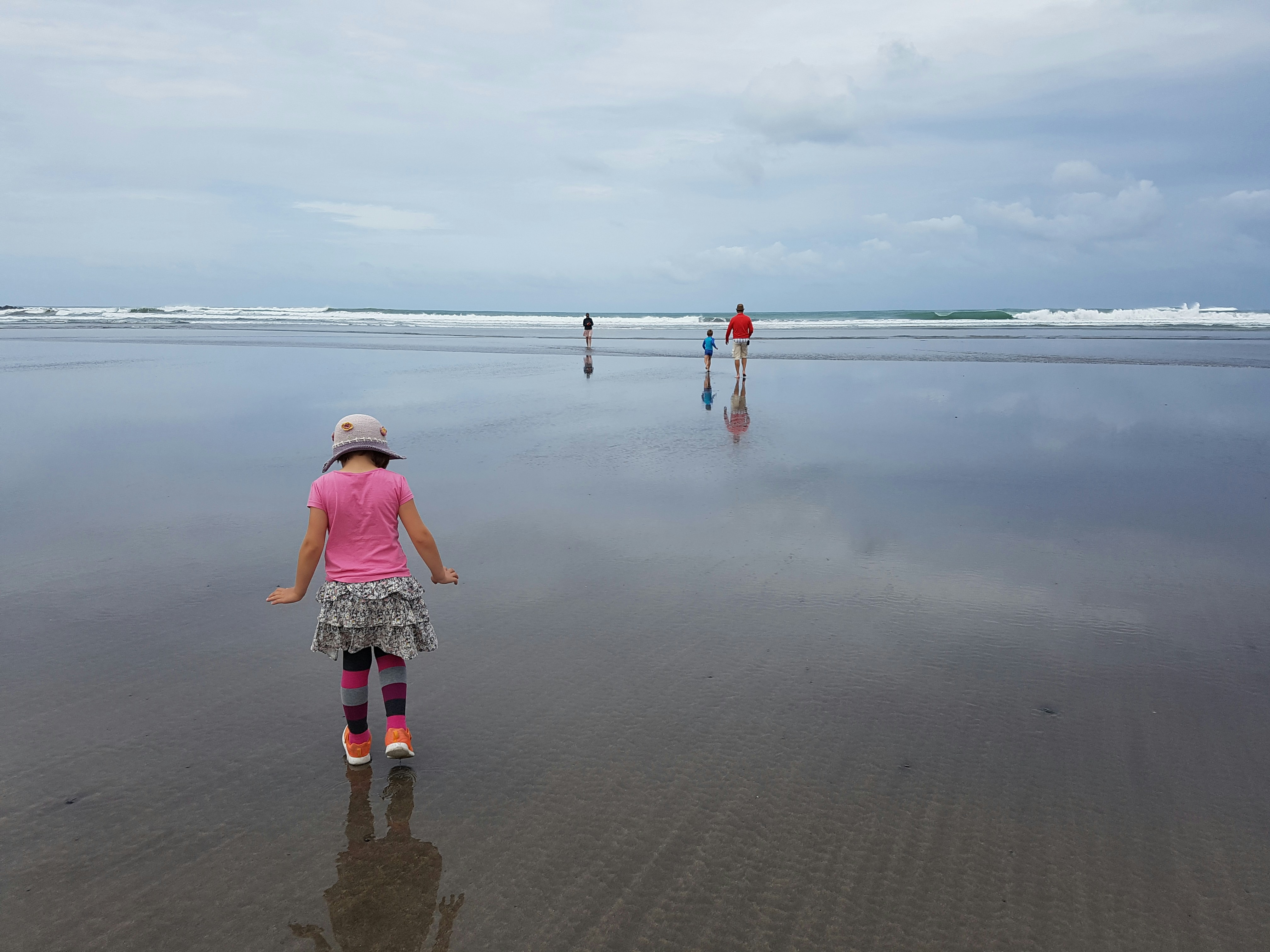 a group of people are walking towards the ocean over sand which is covered by a thin layer of water creating a mirrored surface | Girl in pink shirt runs on wet beach toward ocean