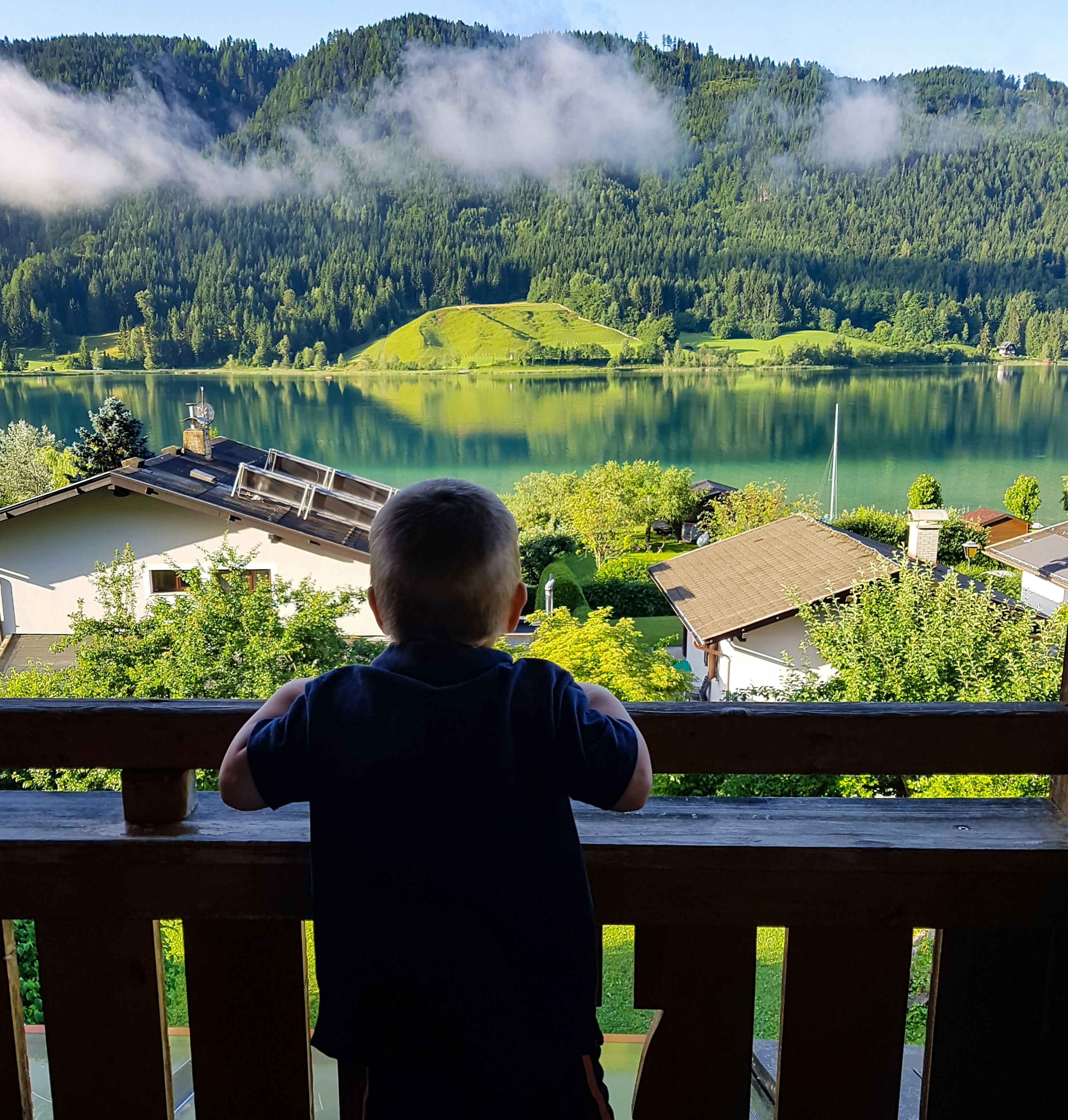 A small boy is enjoying the view over a lake from a balcony | Boy looking at lake and mountains from balcony